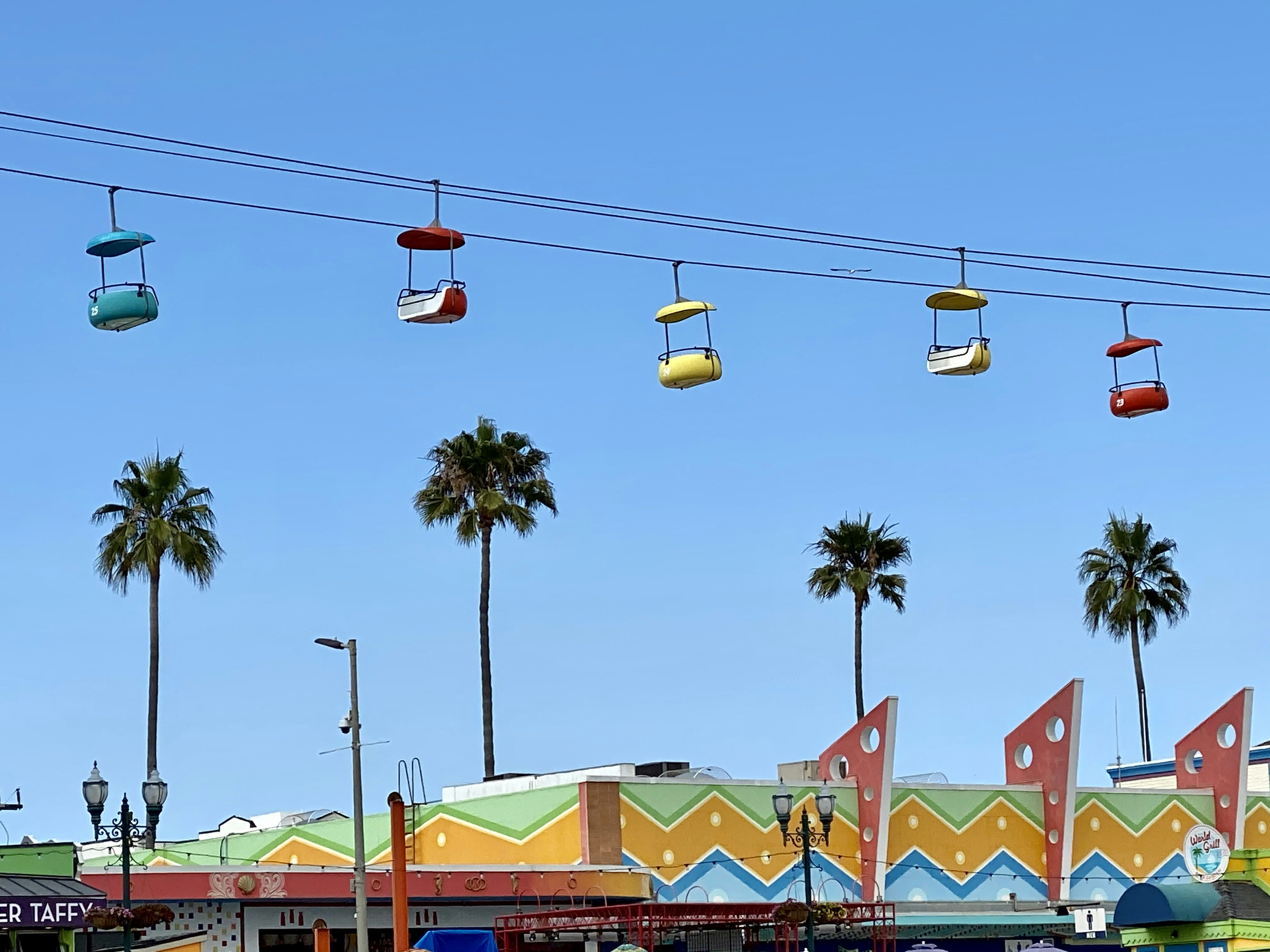 Colorful gondolas suspended from cables above a lively boardwalk adorned with palm trees and bright murals.