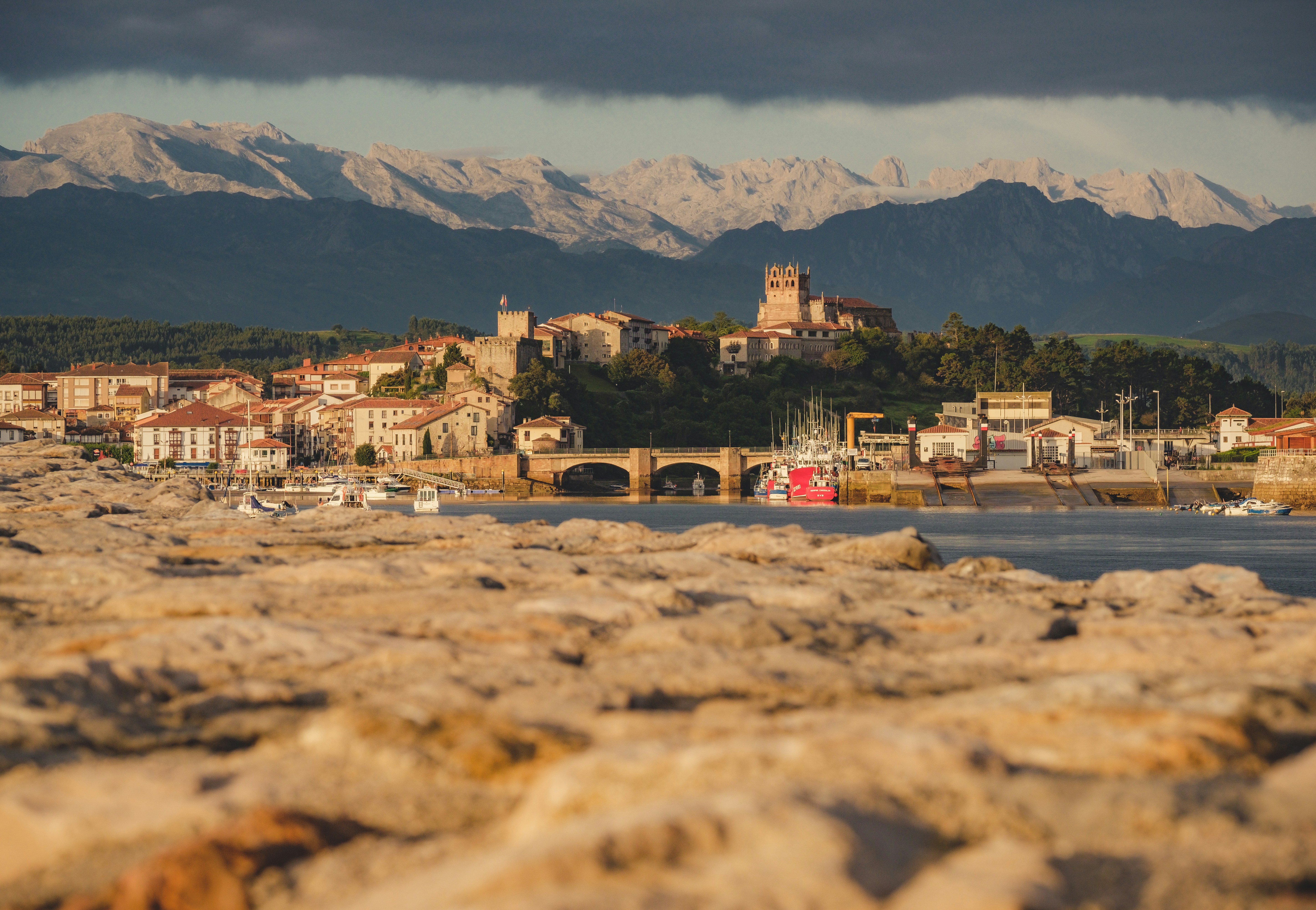 Coastal town with stone buildings and a historic castle set against a backdrop of snow-capped mountains.