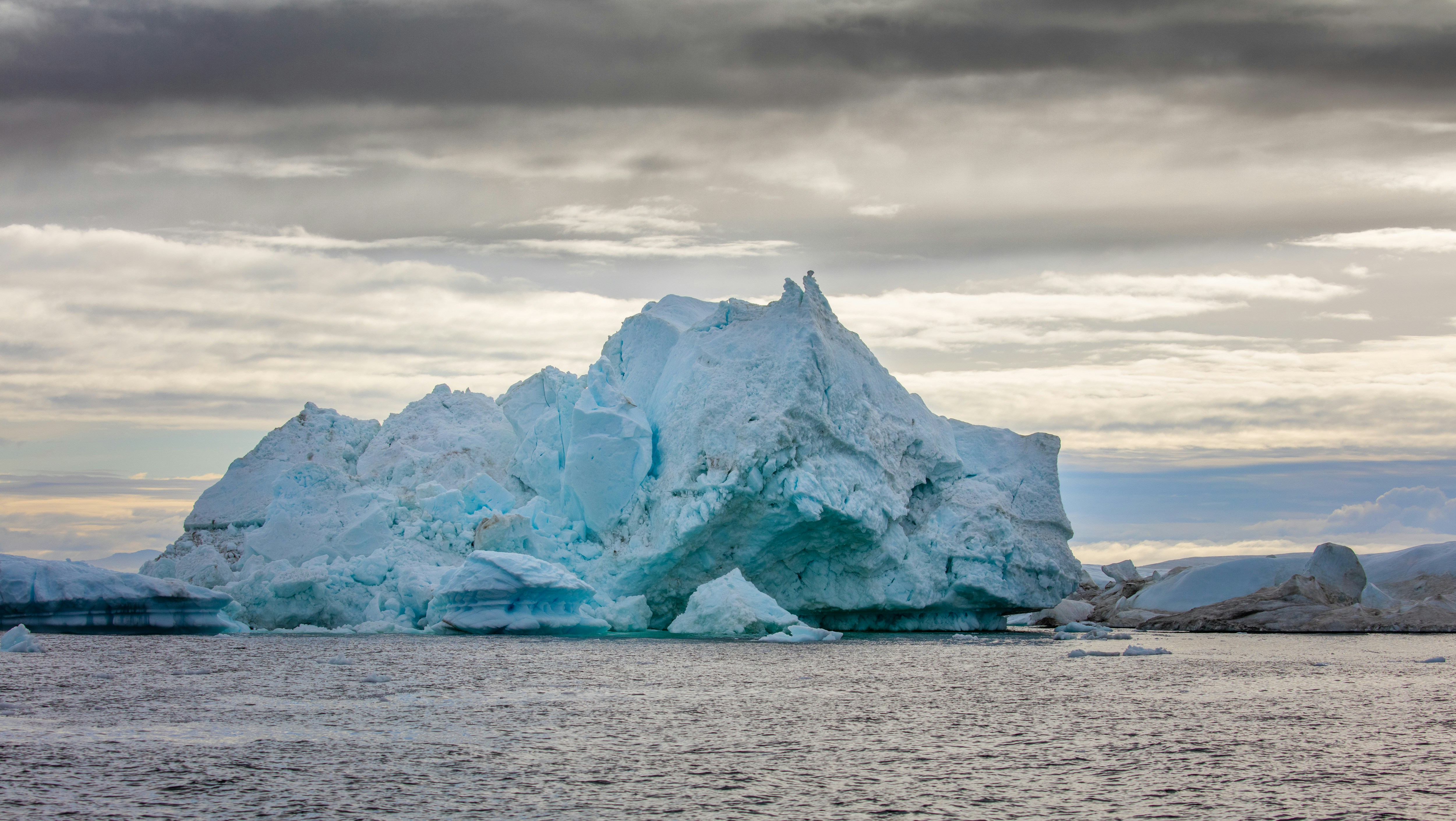 This captivating image showcases a massive iceberg floating serenely under a dramatic, cloud-laden sky. The iceberg's intricate textures and shades of icy blue and white contrast beautifully with the muted grays of the overcast atmosphere, creating a striking visual harmony. The composition emphasizes the iceberg's grandeur and the subtle play of light and shadow, evoking a sense of awe and tranquility.