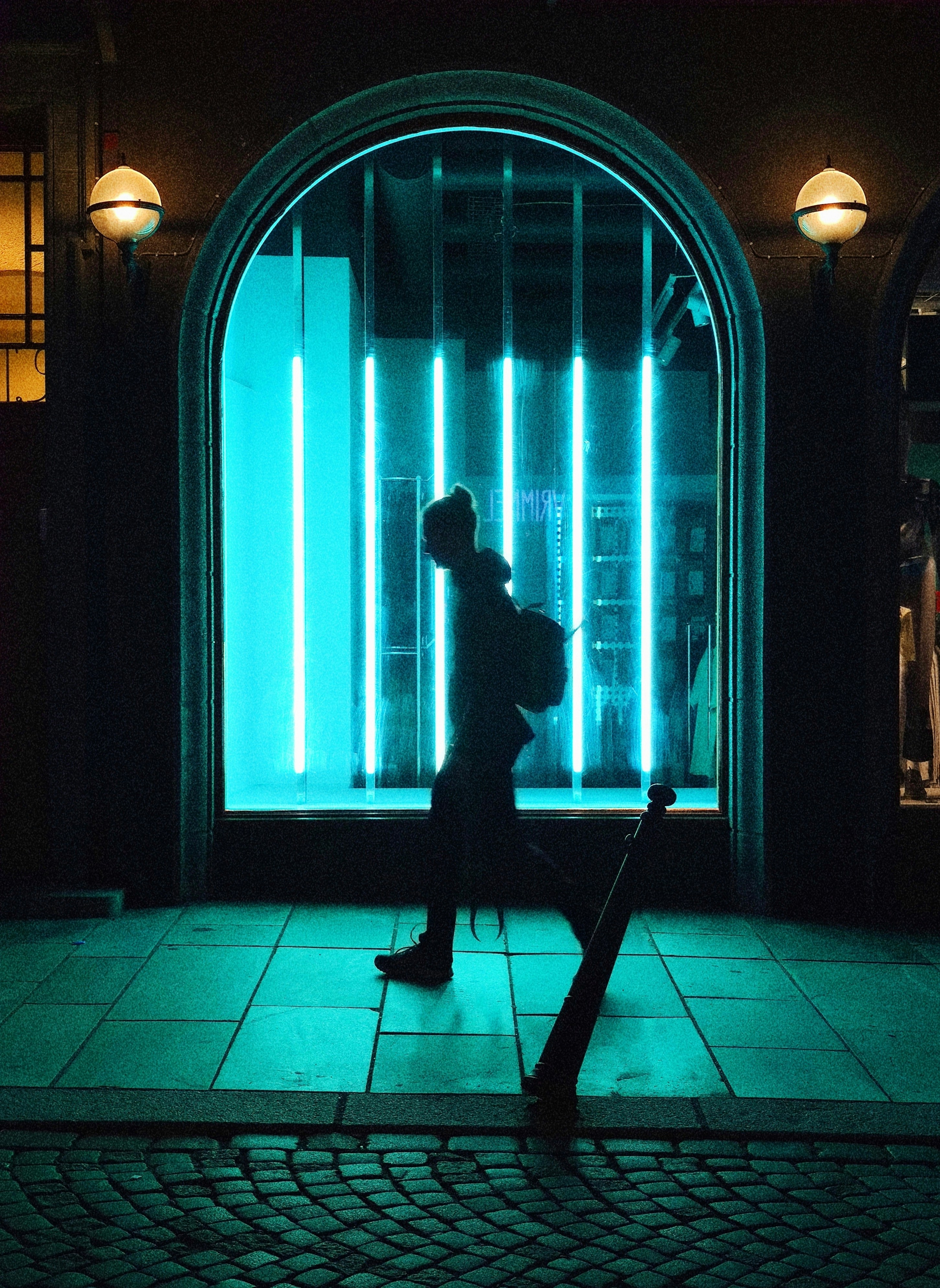 Silhouette of a person walking past a storefront illuminated by striking blue neon lights. The contrast between light and shadow creates a dynamic urban scene.