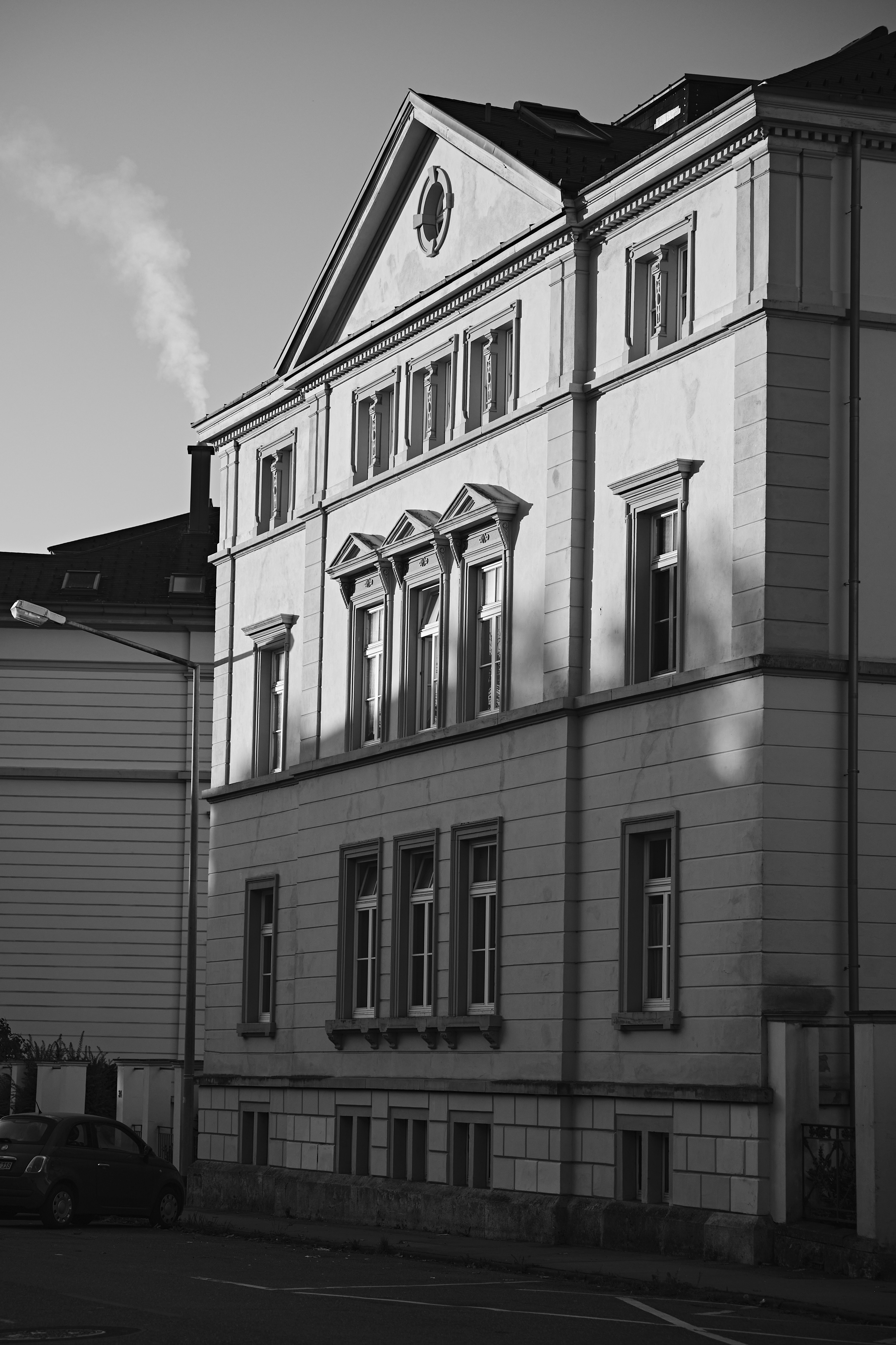 Historic building facade illuminated by soft light, showcasing intricate window designs and shadows. The structure stands in stark contrast against its modern surroundings.