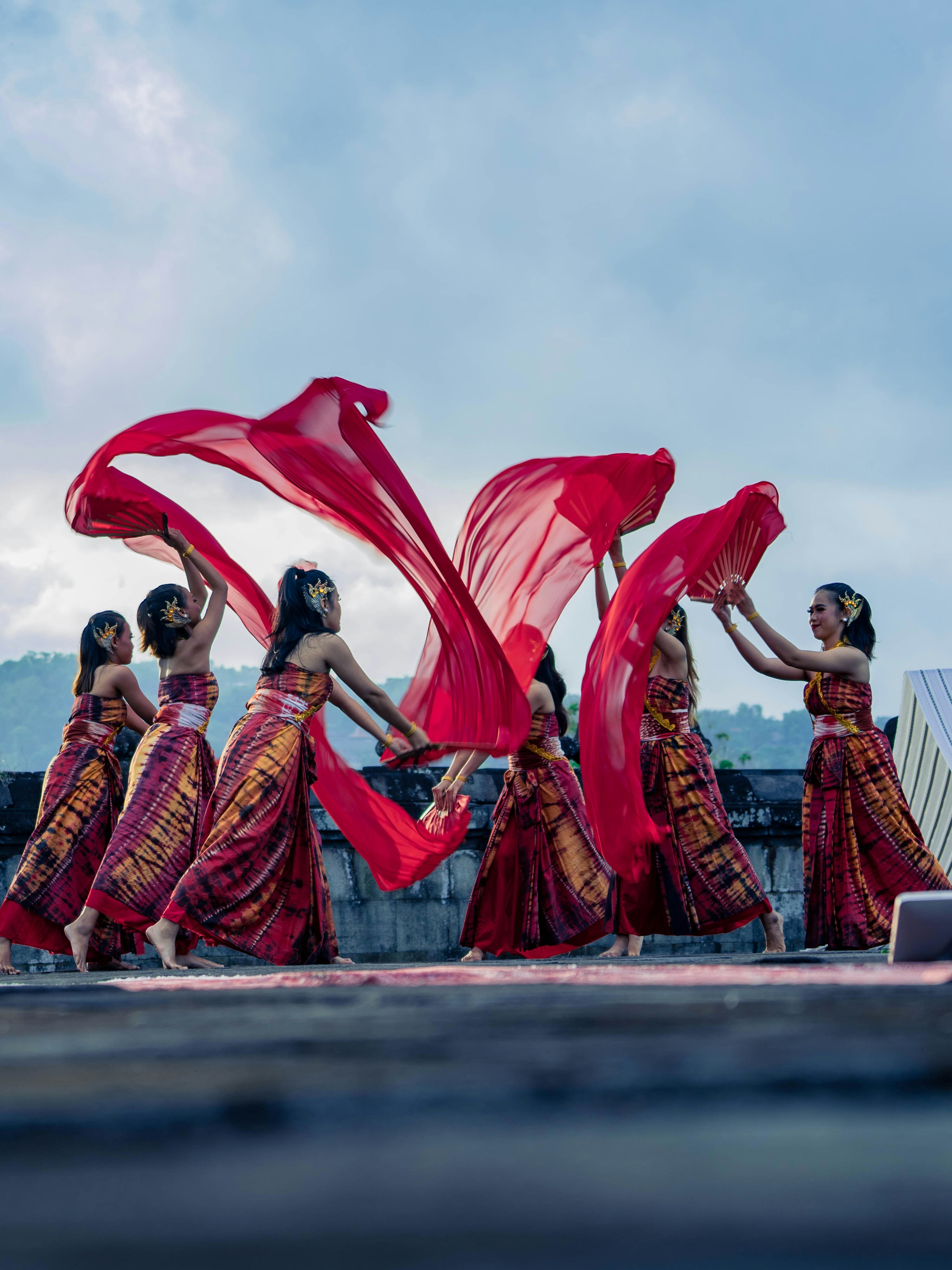 Six dancers in vibrant attire gracefully manipulate flowing red fabrics against a scenic backdrop, embodying cultural heritage through movement.