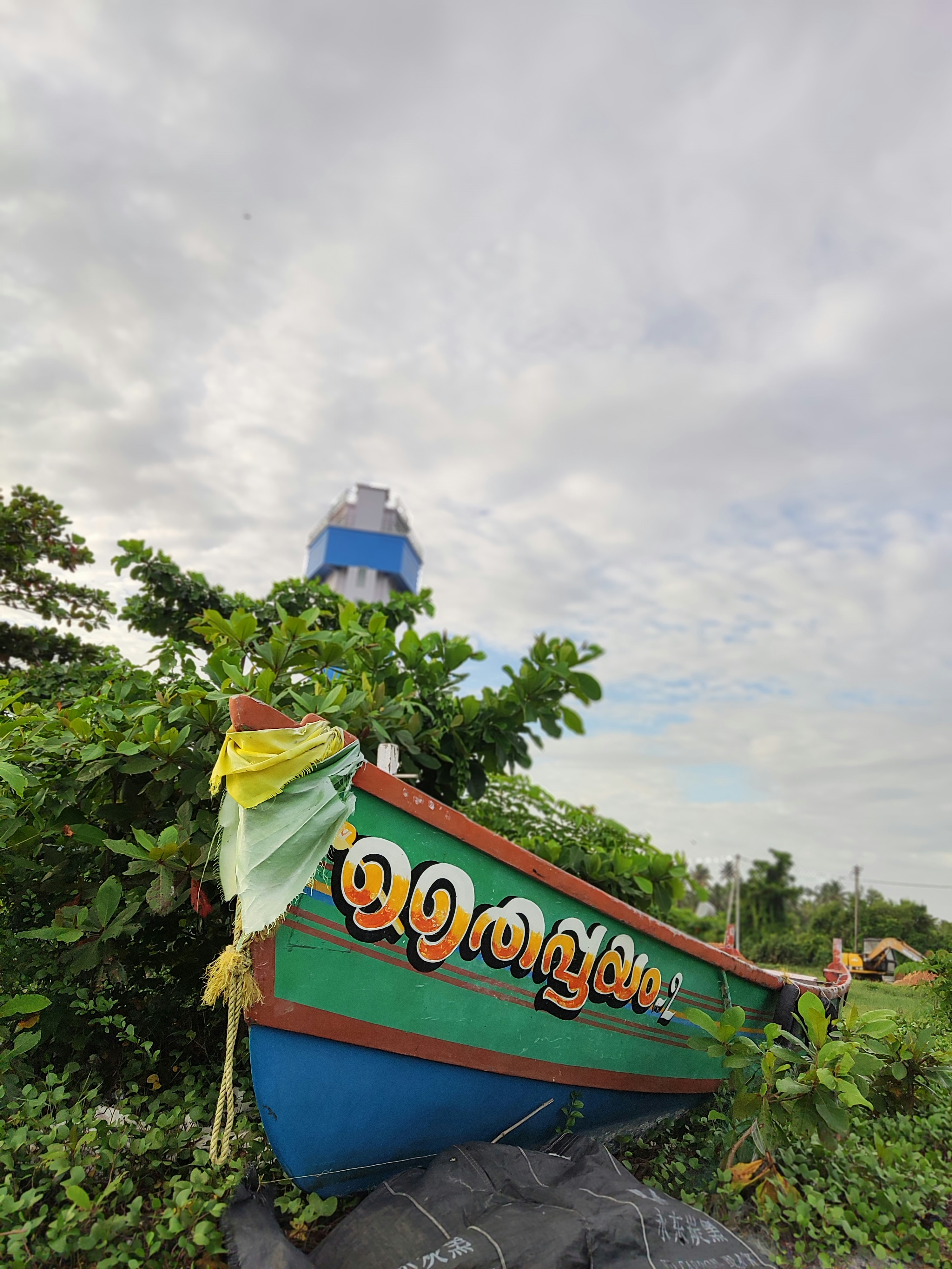 Colorful wooden boat resting on overgrown grass beneath a cloudy sky, with a multicolor cloth draped over the bow.