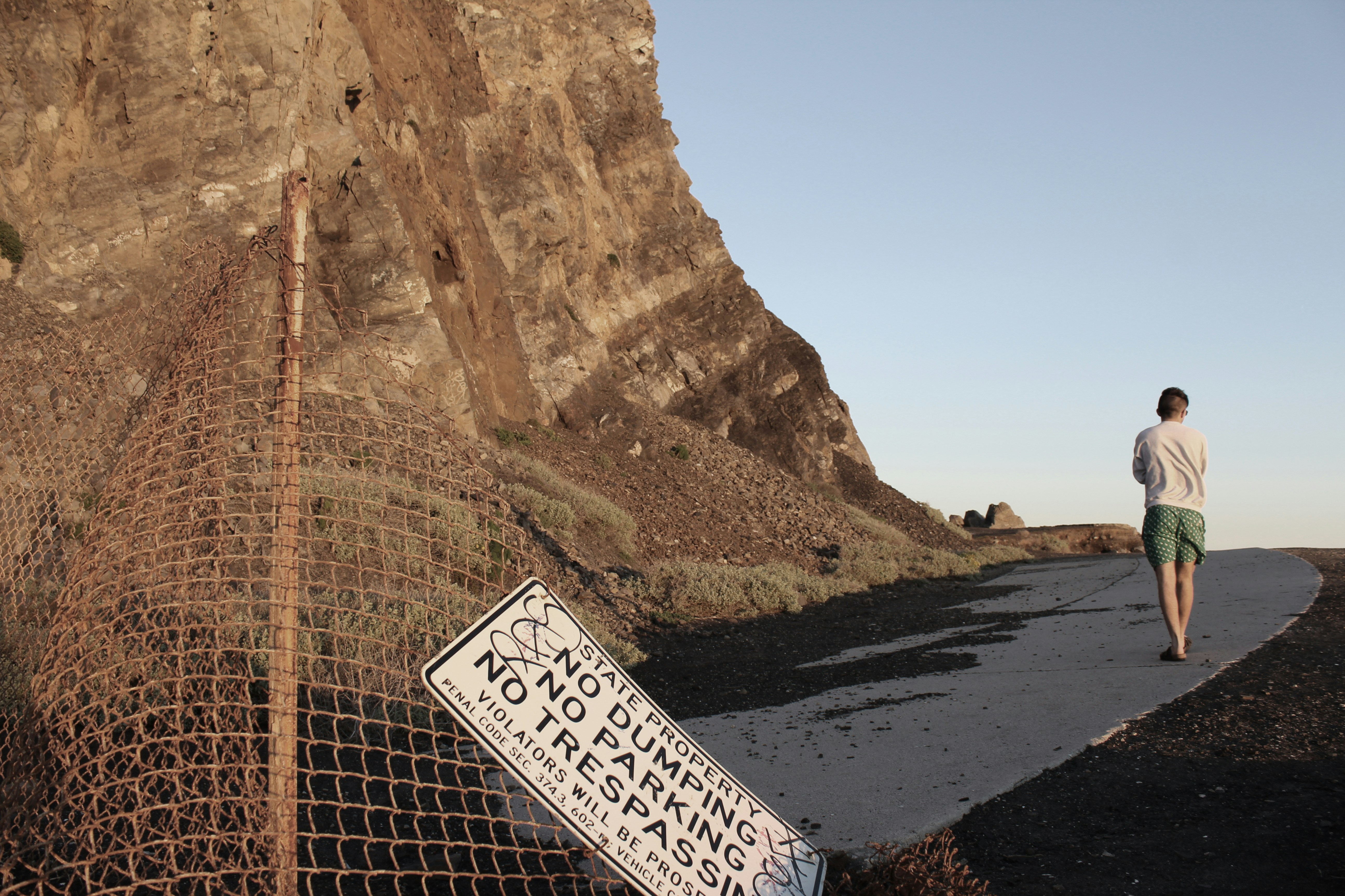 A solitary figure walks along a weathered path, bordered by a rusted fence and a warning sign, against the backdrop of a towering cliff. 