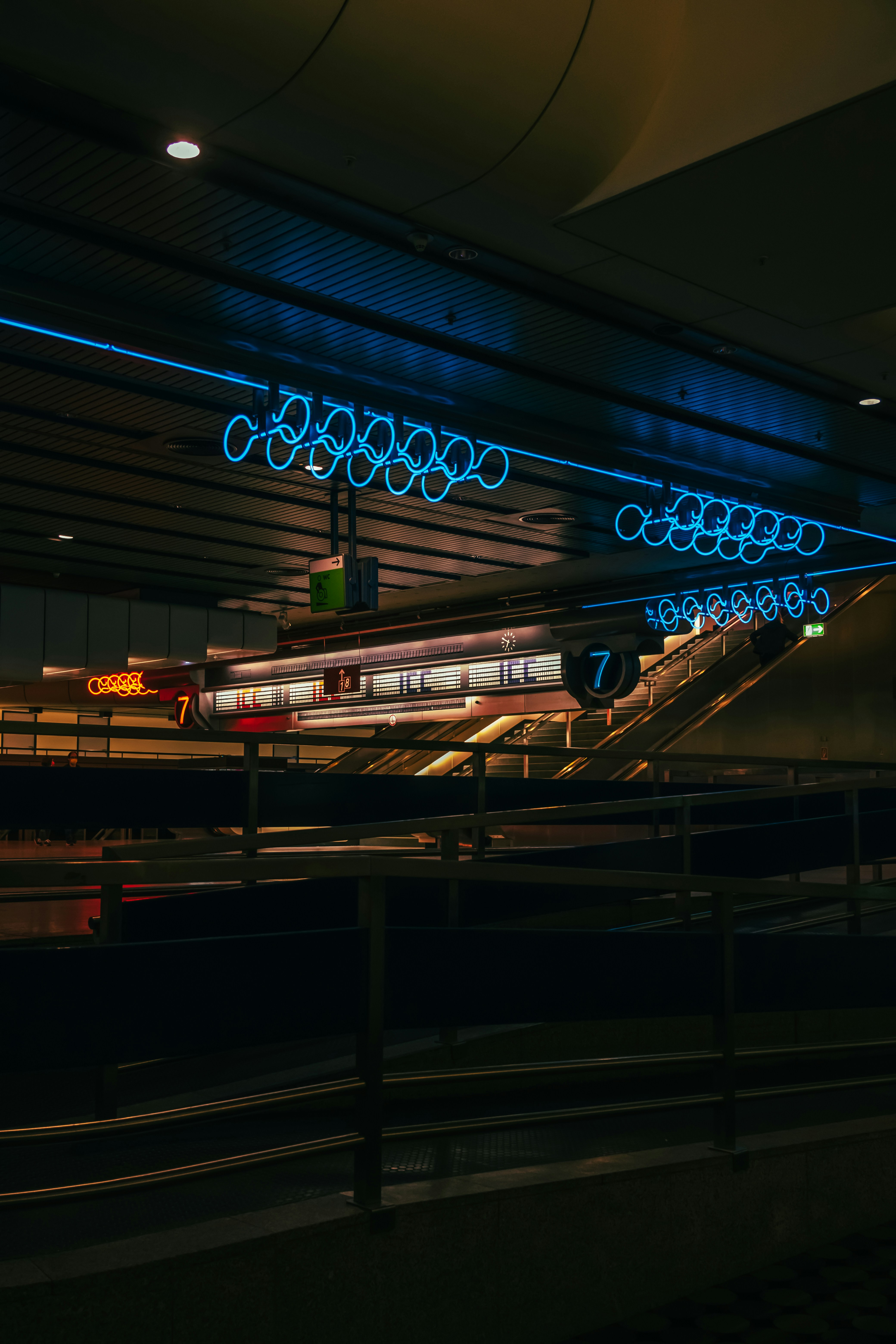Illuminated signage and neon lights guide the way in a modern transit hub, creating a striking visual contrast against the dim surroundings.