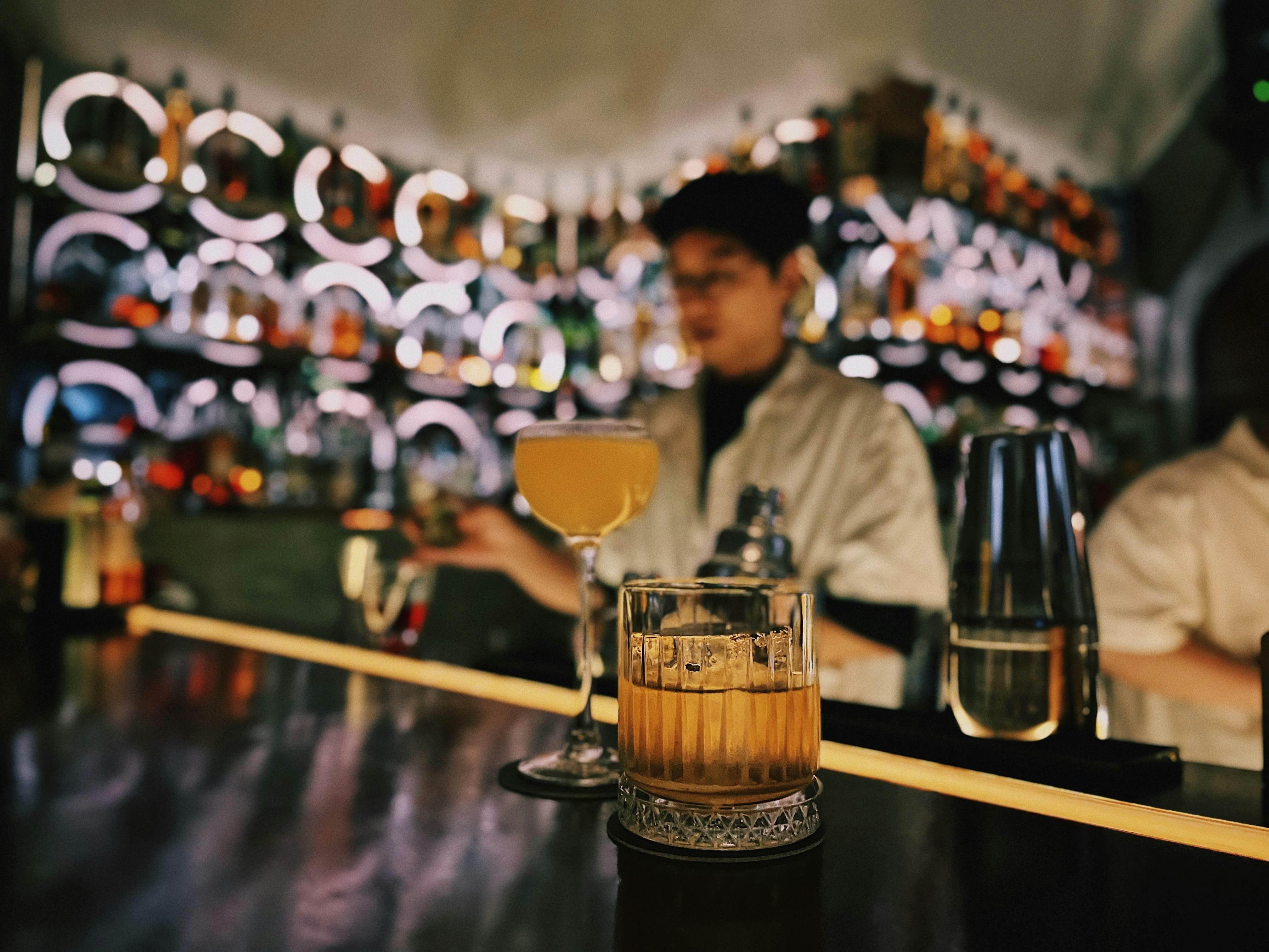 A close-up of two cocktails on a bar counter, with a bartender skillfully preparing drinks in the background. The vibrant bar shelves are illuminated with colorful bottles.