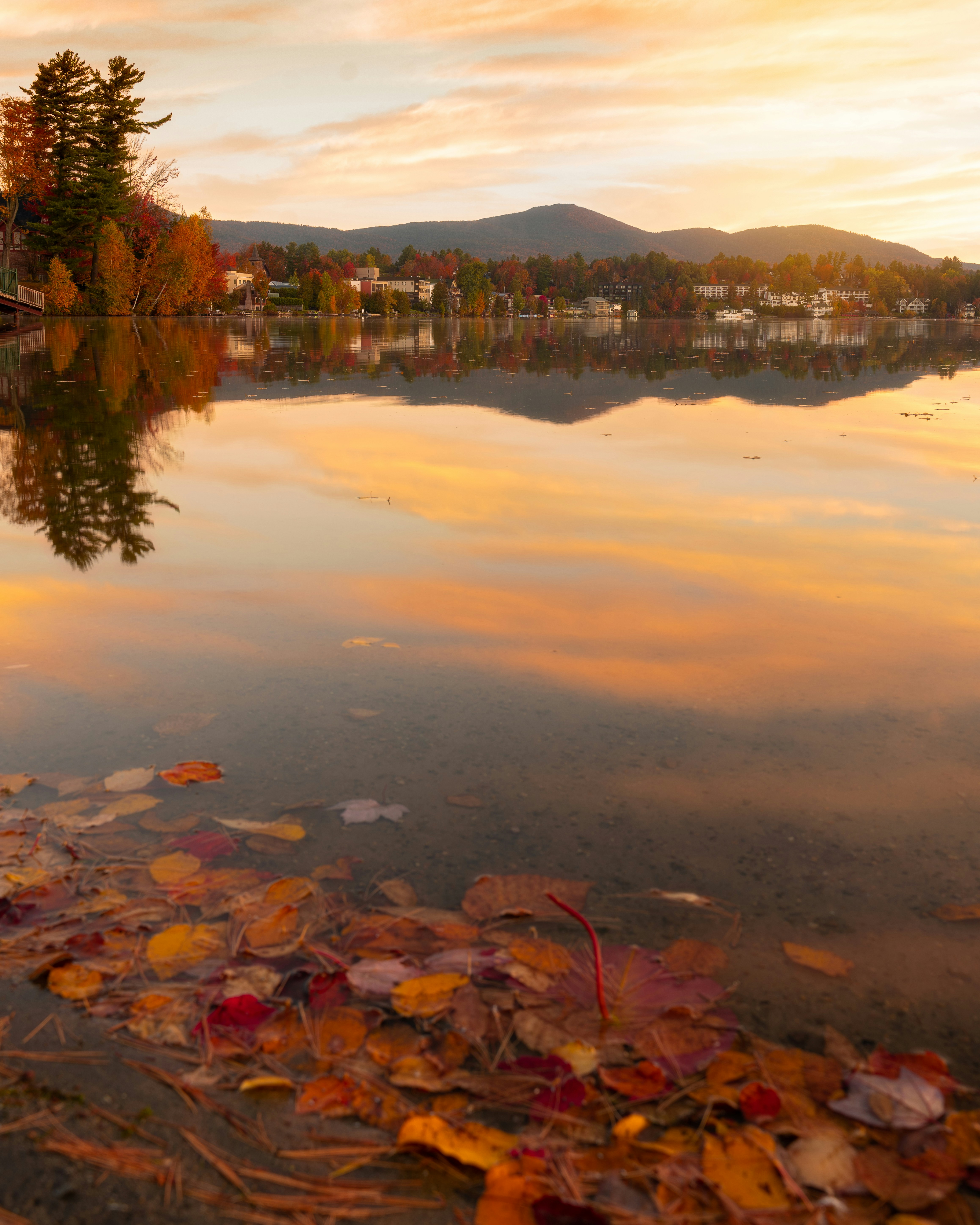 Colorful autumn leaves float on the lake's surface, reflecting the vibrant hues of the sunset and surrounding trees.