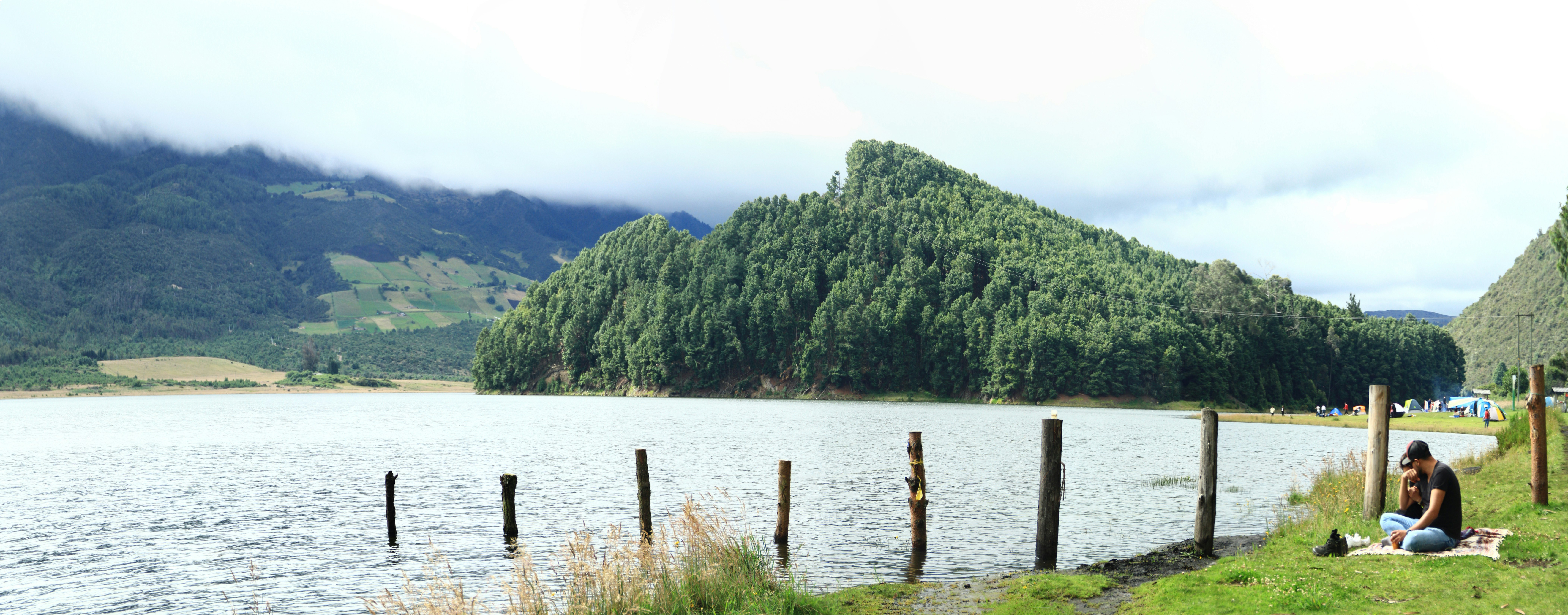 Person sitting on a blanket beside a serene lake with lush green hills under a cloudy sky.