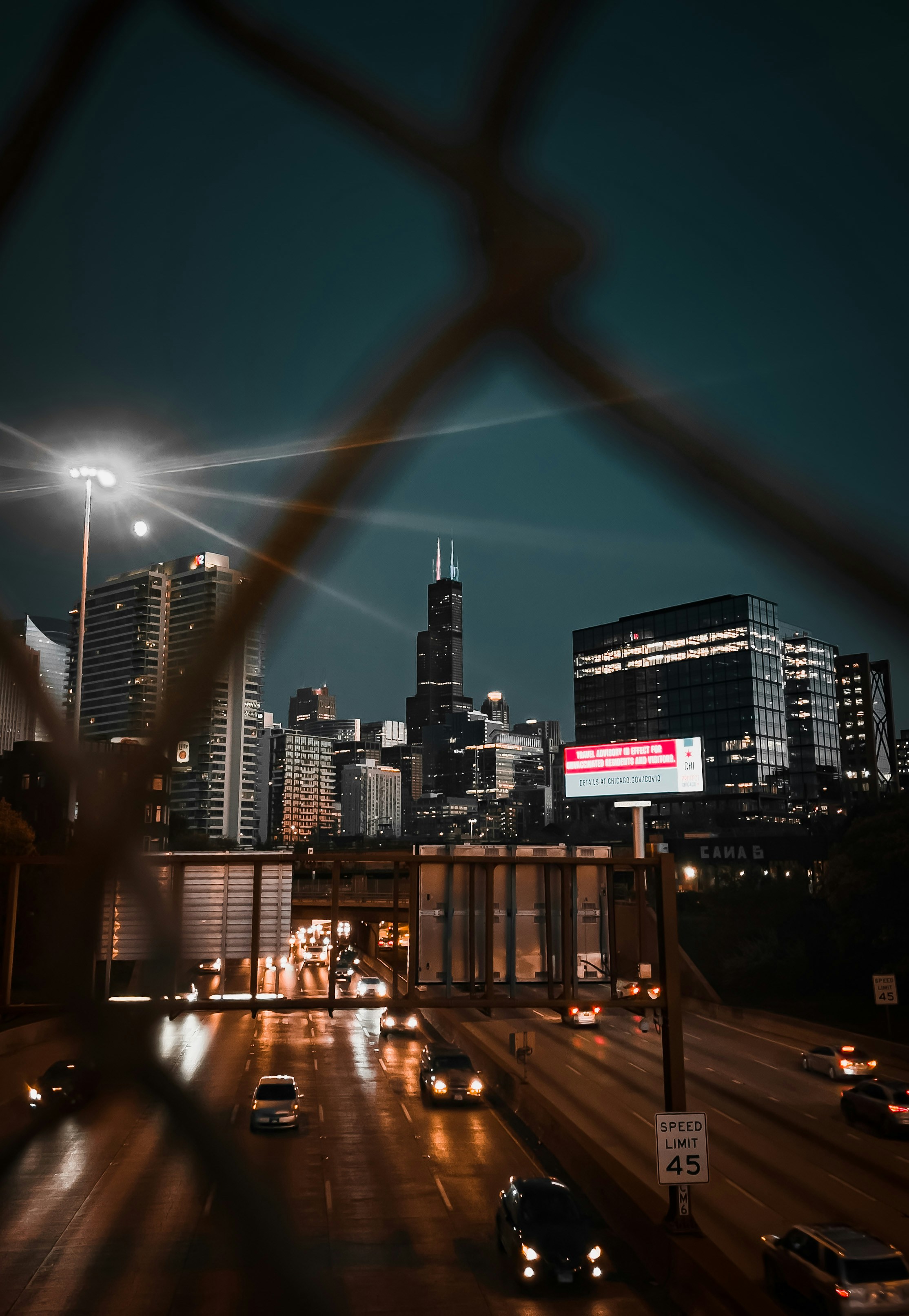 City skyline illuminated at night, viewed through a fence, showcasing the interplay of light and architecture. A highway runs below, filled with moving vehicles.
