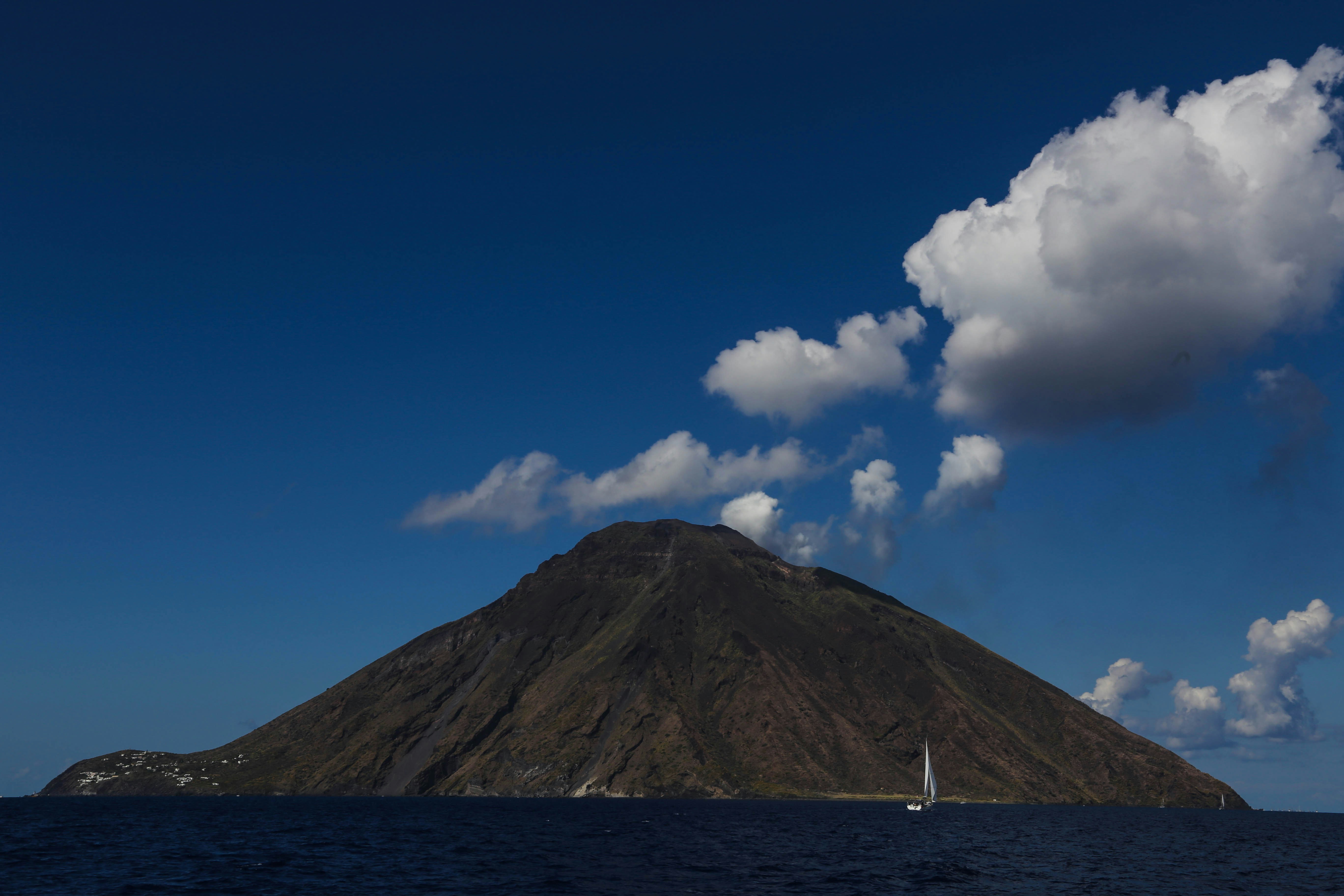 Sailboat gliding past the towering Stromboli volcano under a sky dotted with fluffy clouds.