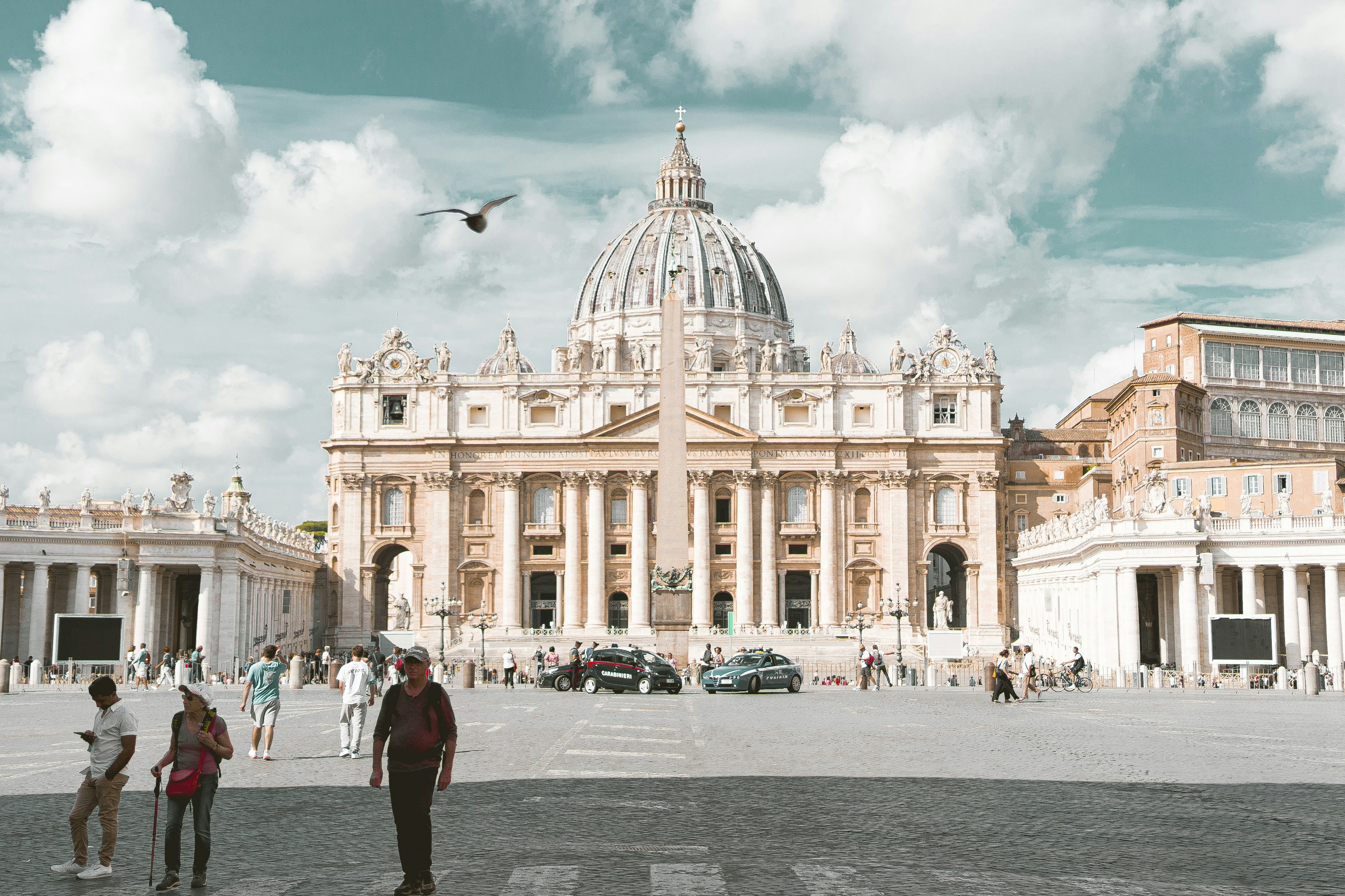 St. Peter's Basilica stands majestically under a blue sky, surrounded by visitors exploring the iconic square. A bird soars gracefully above the historic architecture.