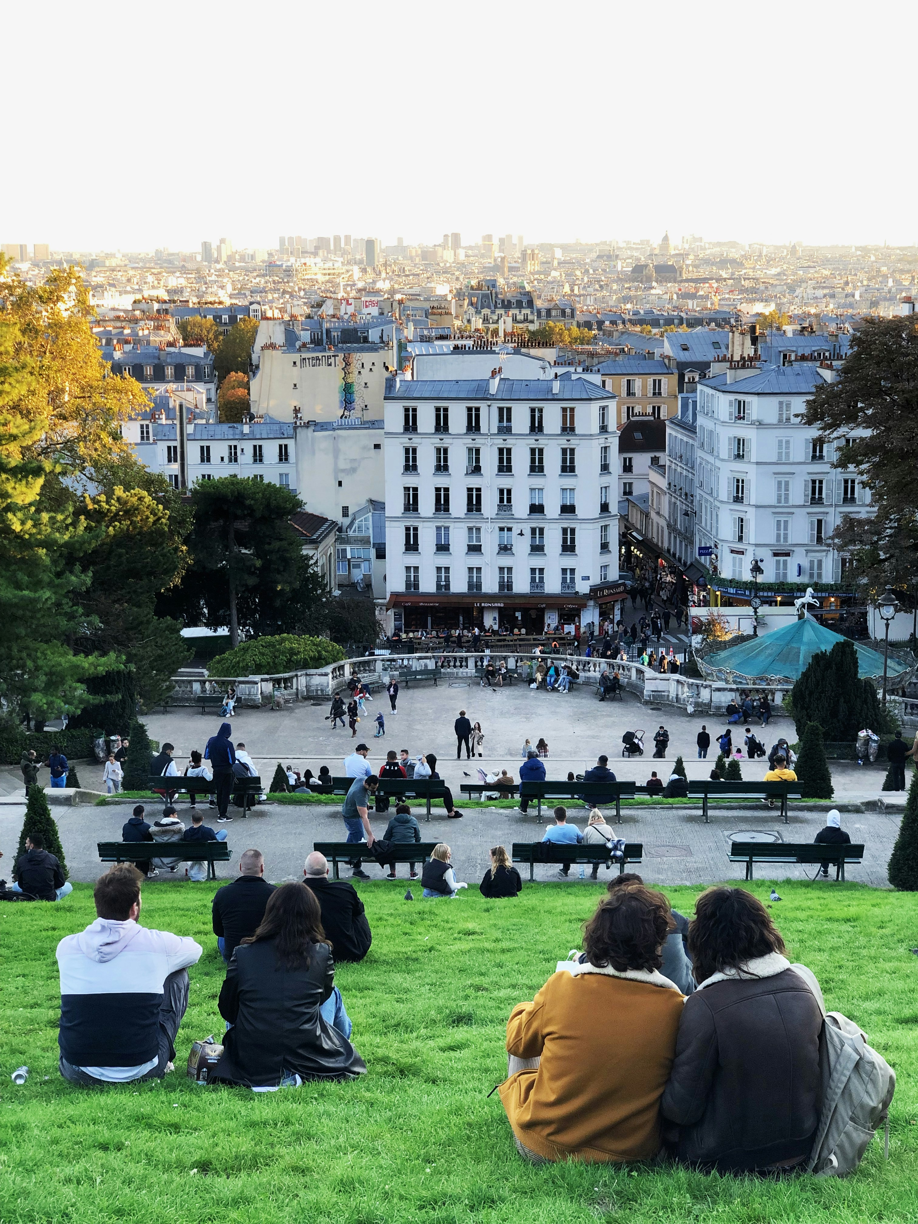 Couple sitting on a grassy hill overlooking a bustling urban plaza filled with people, framed by historic buildings and distant city skyline.