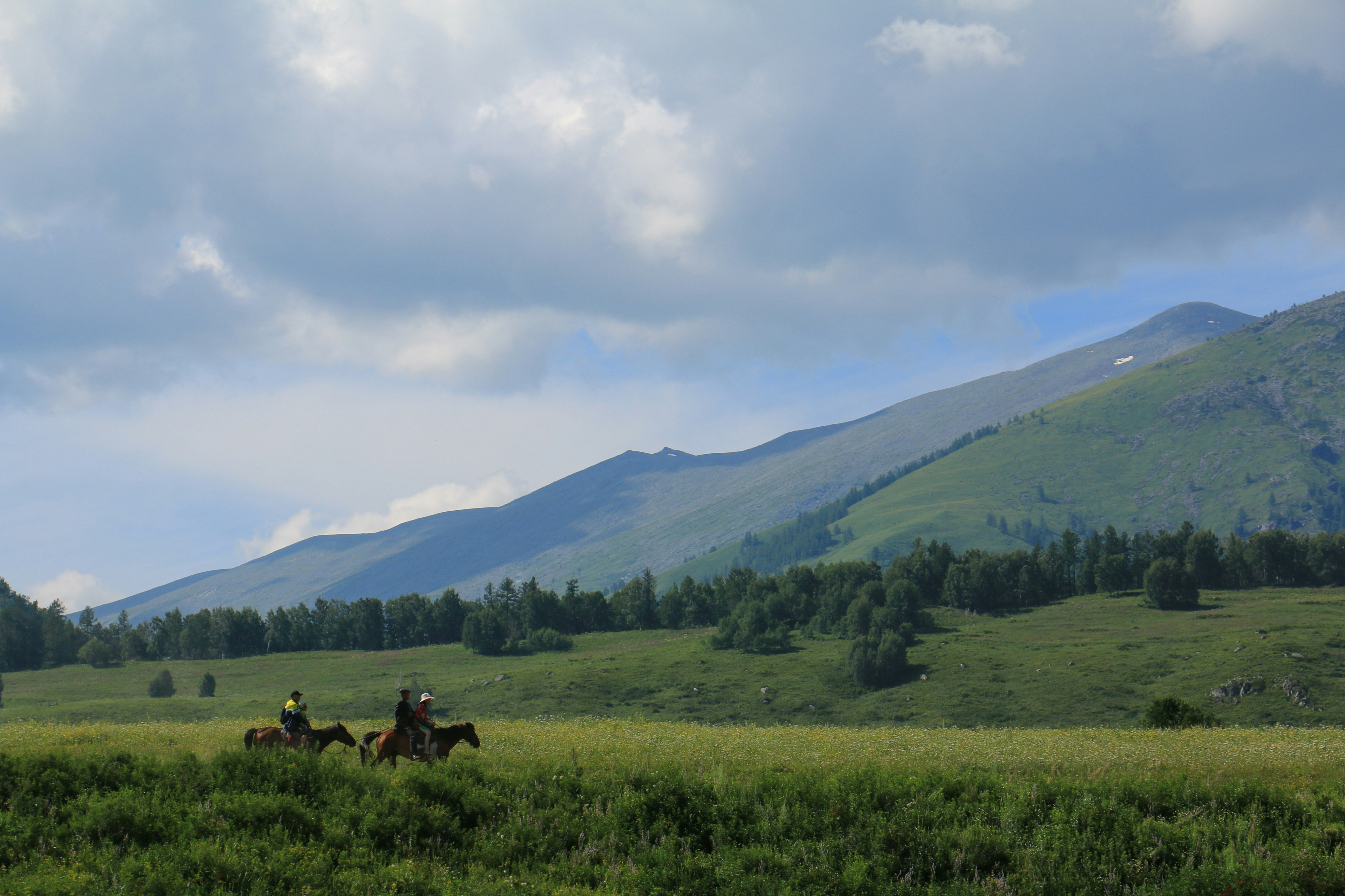 Two riders on horseback traverse a lush green meadow with rolling hills and distant mountains under a partly cloudy sky.