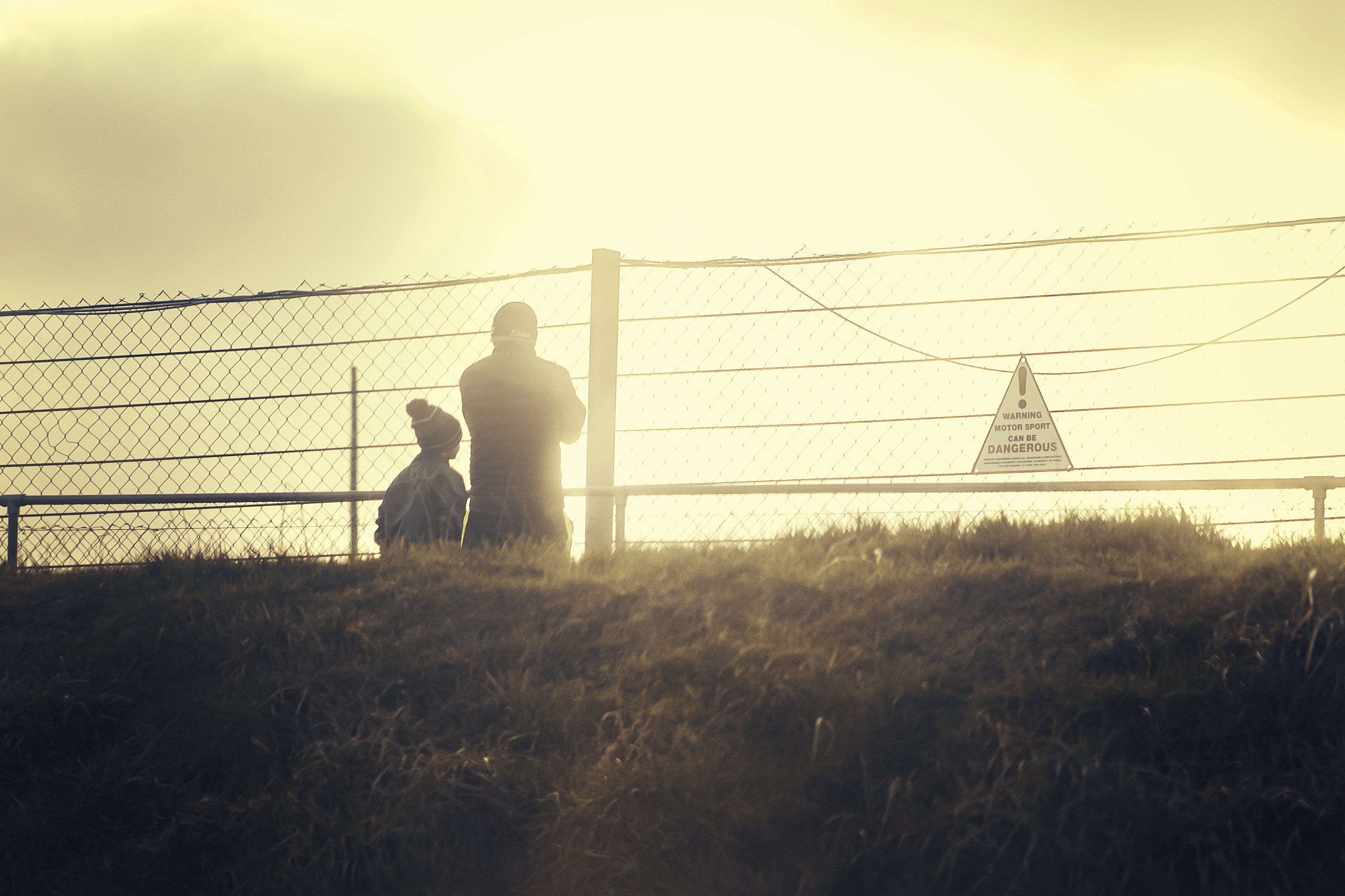 Two figures stand silhouetted against a soft, glowing horizon, framed by a fence and a caution sign. The scene evokes a sense of contemplation and quietude.