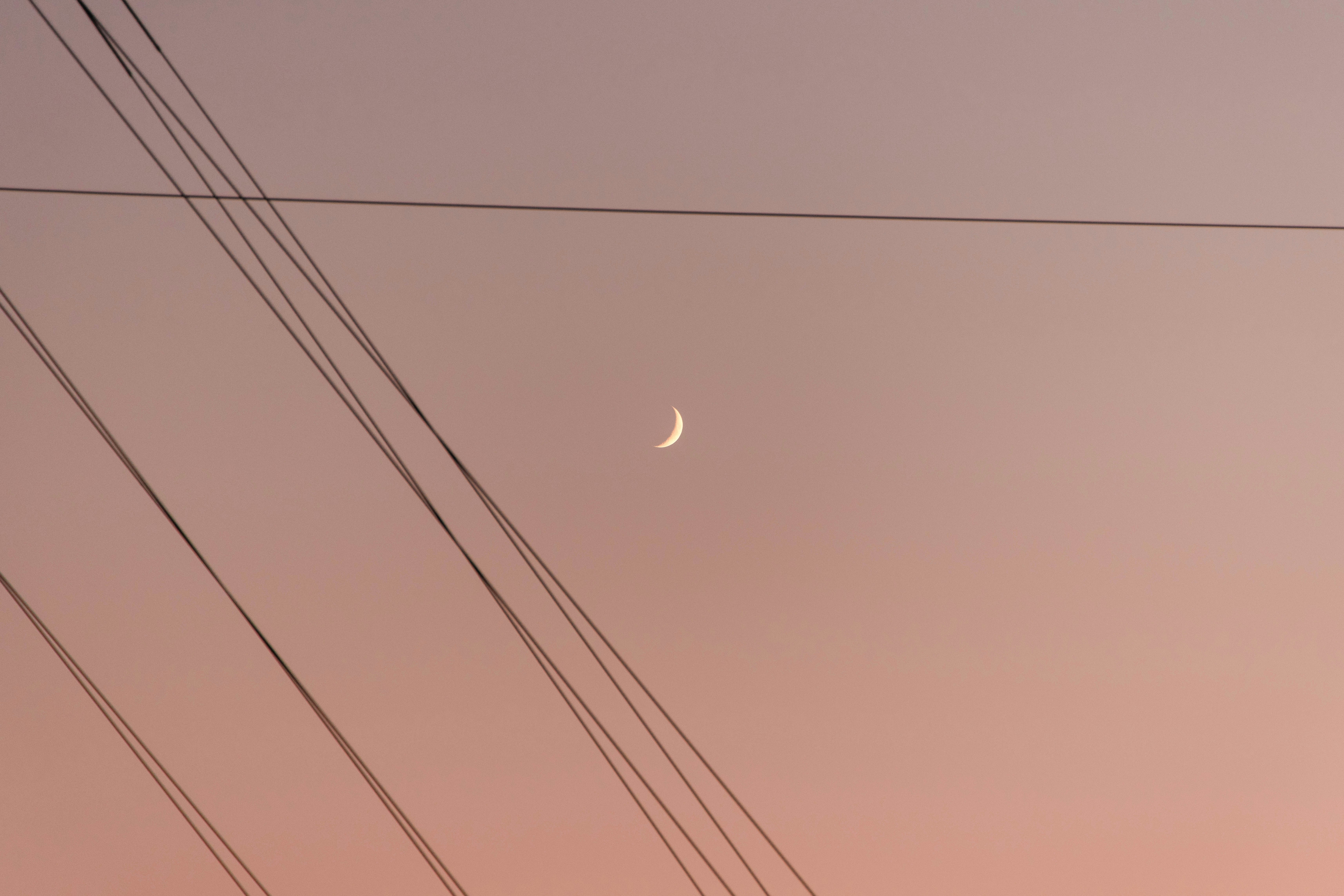 Crescent moon peeks through a soft pastel sky, intersected by power lines at dusk.