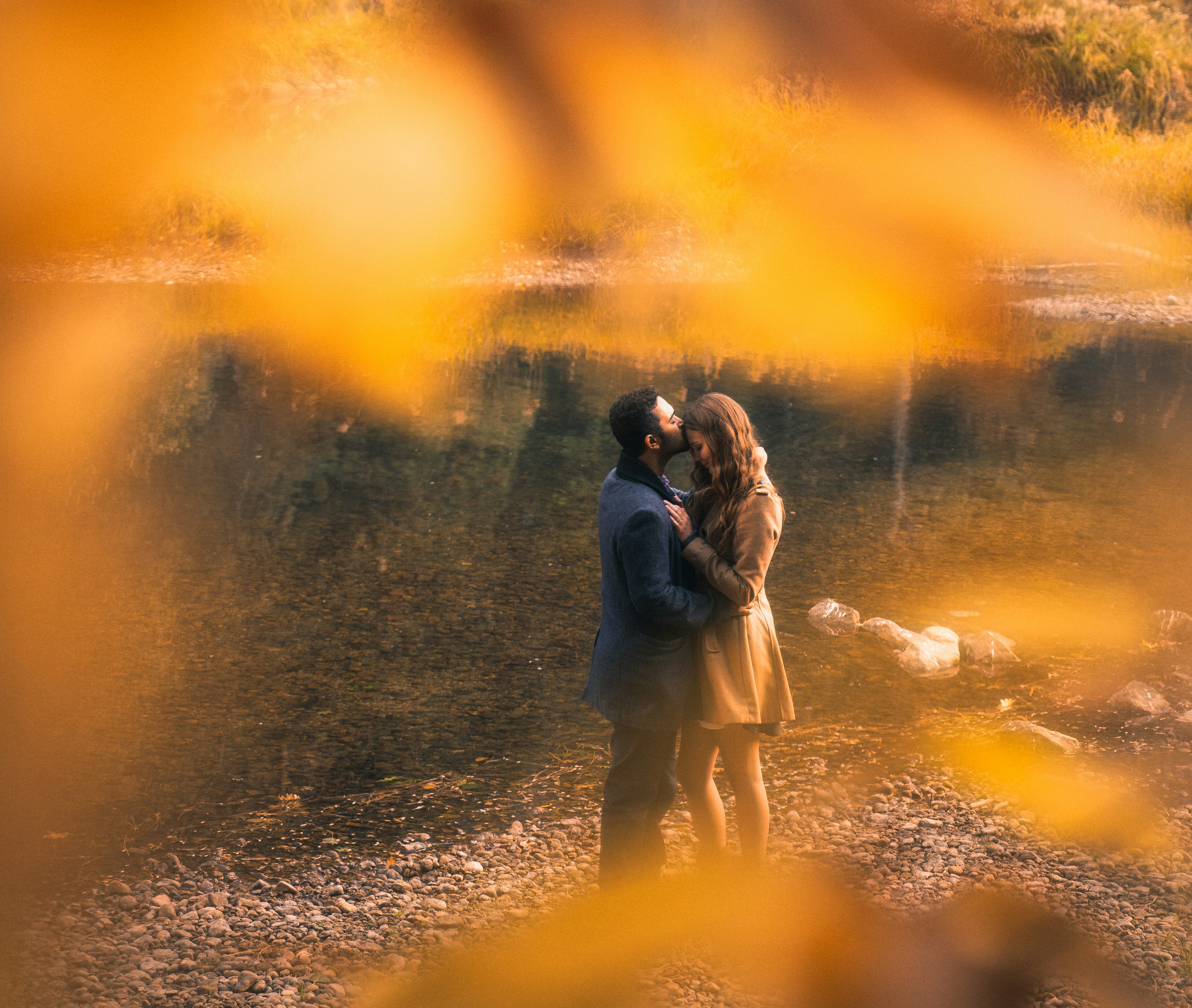 Couple sharing a tender moment by the water's edge, framed by vibrant autumn leaves. The warm colors enhance the intimacy of their connection.