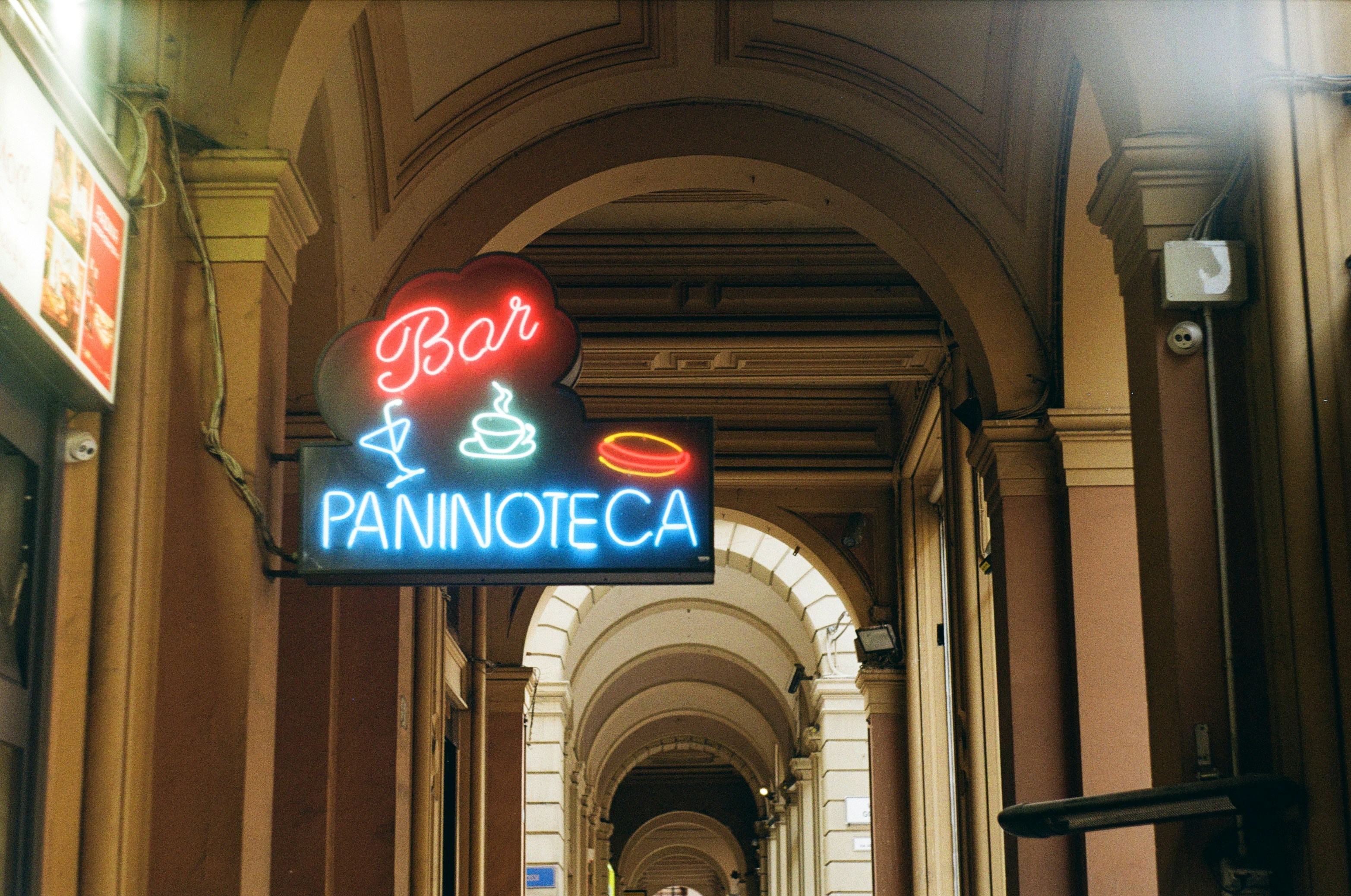 Photograph of a neon Bar Paninoteca sign hanging beside a narrow arched passage, casting colorful glow along brick walls. The repeating arches lead the eye toward the deeper corridor.