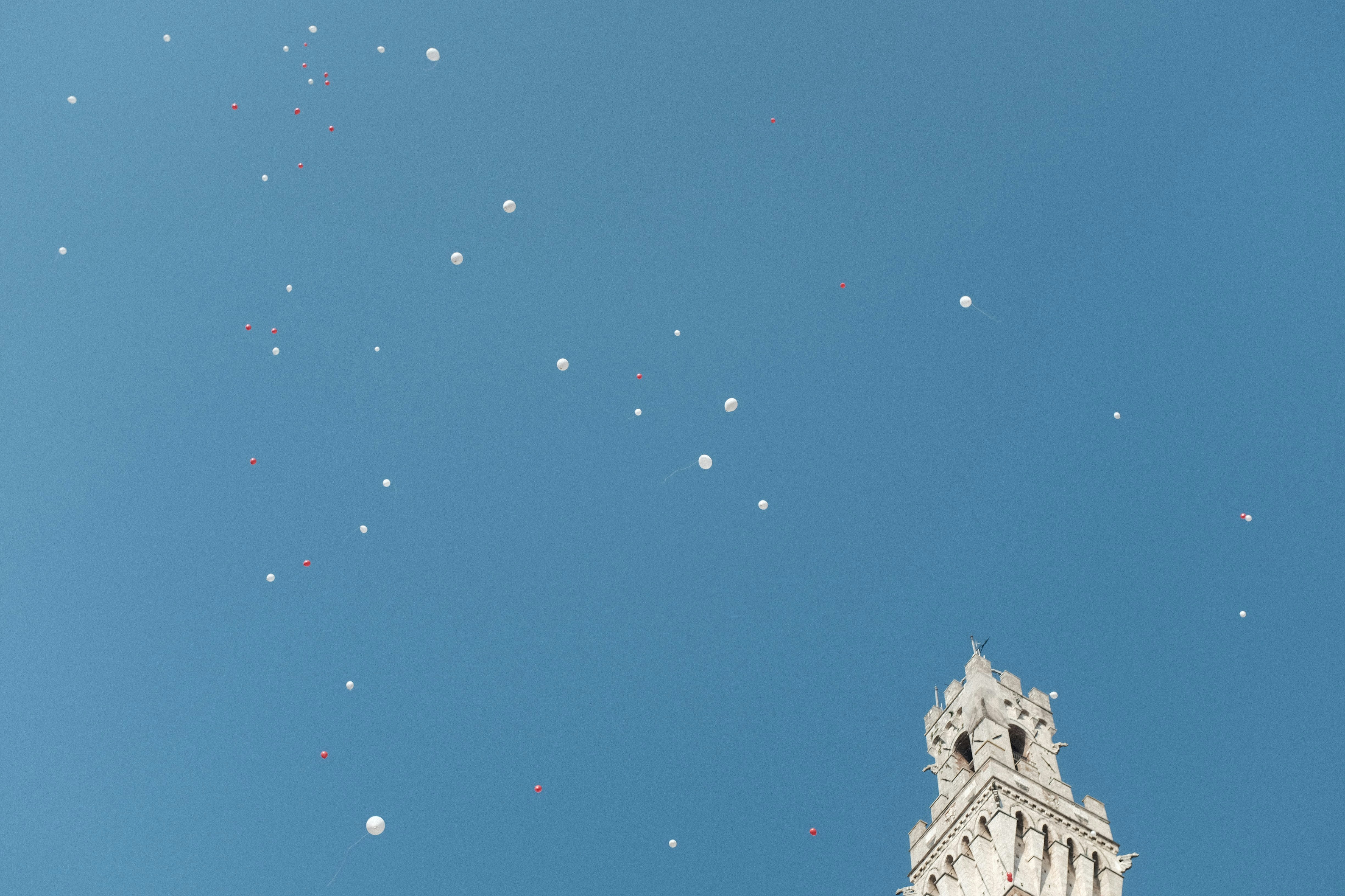 White balloons drift above a detailed tower against a clear blue sky.