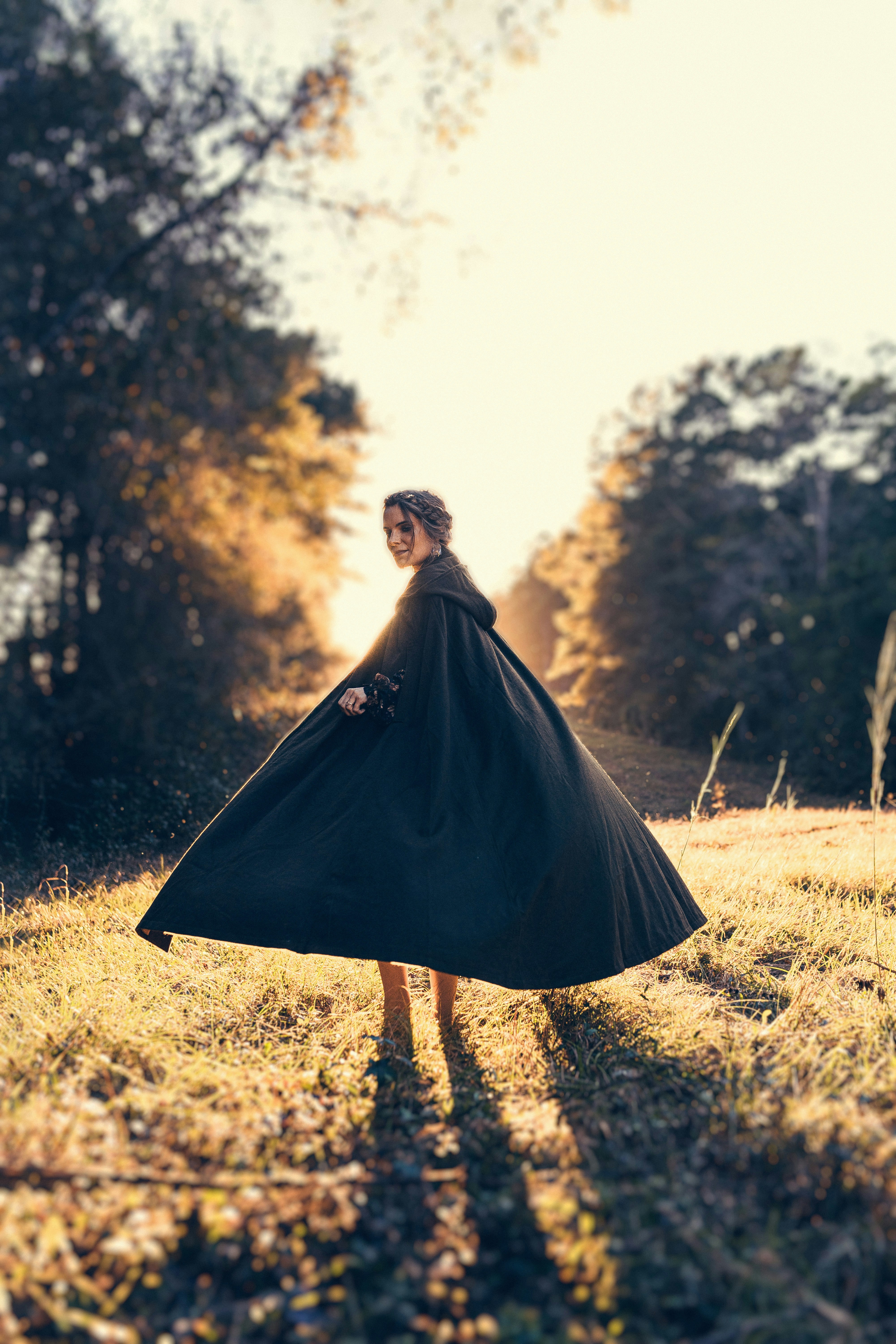 Woman in a flowing black cloak twirls barefoot in a sunlit field surrounded by autumn foliage. The warm light enhances the serene atmosphere.