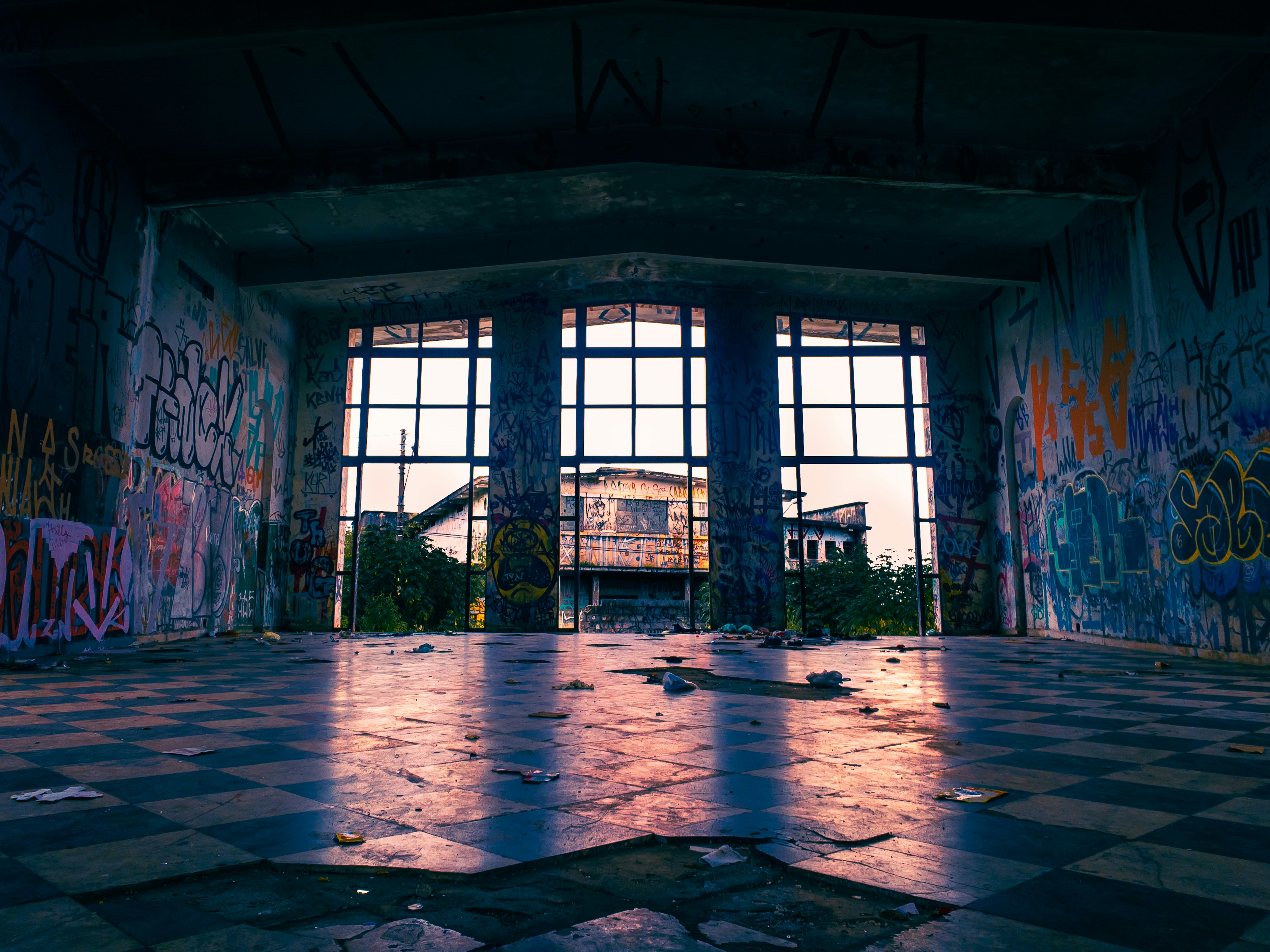An abandoned room with large windows revealing a view of overgrown vegetation and graffiti-covered walls. The floor is scattered with debris, highlighting the decay.