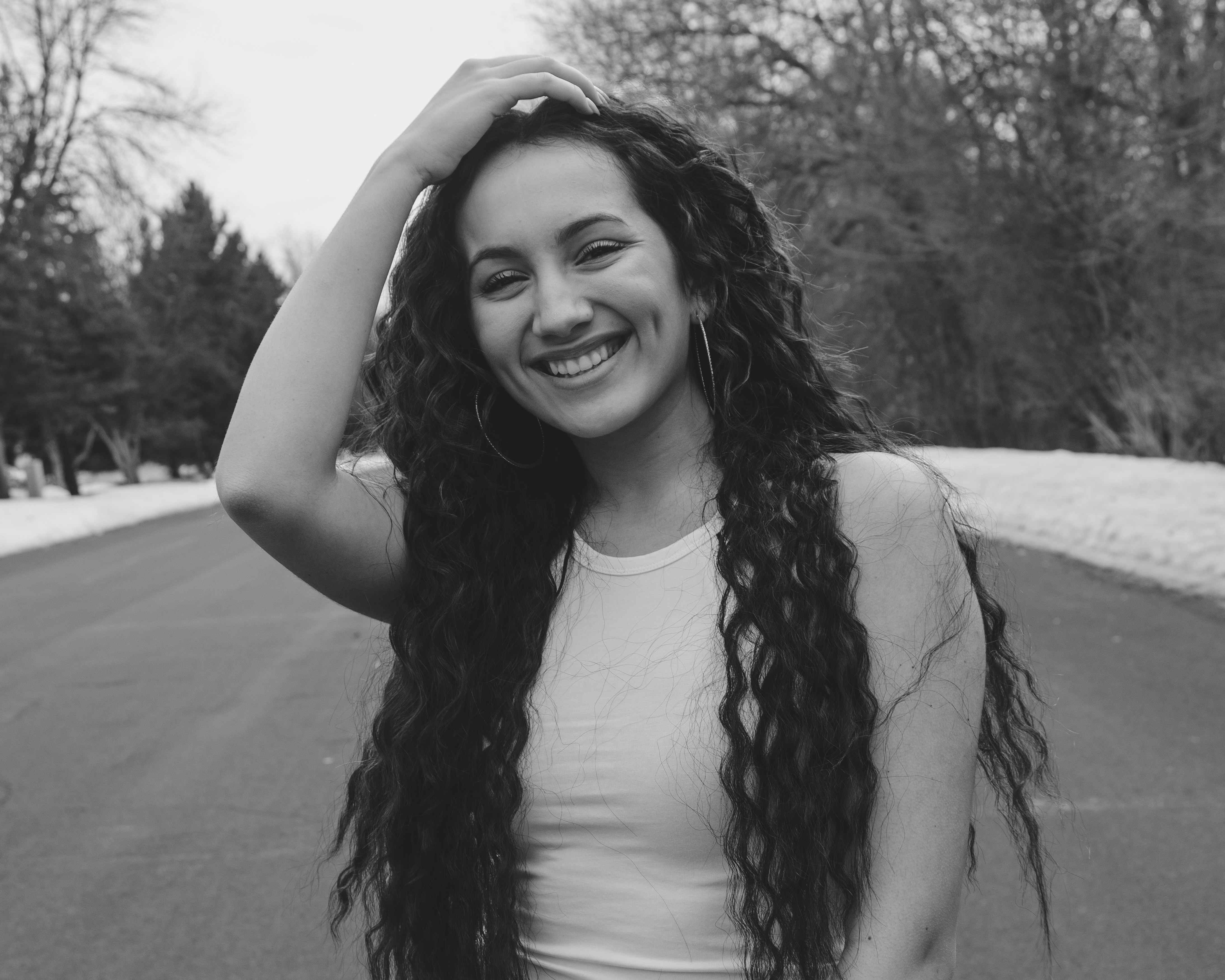 Smiling woman with long curly hair stands on a snowy road, surrounded by bare trees.