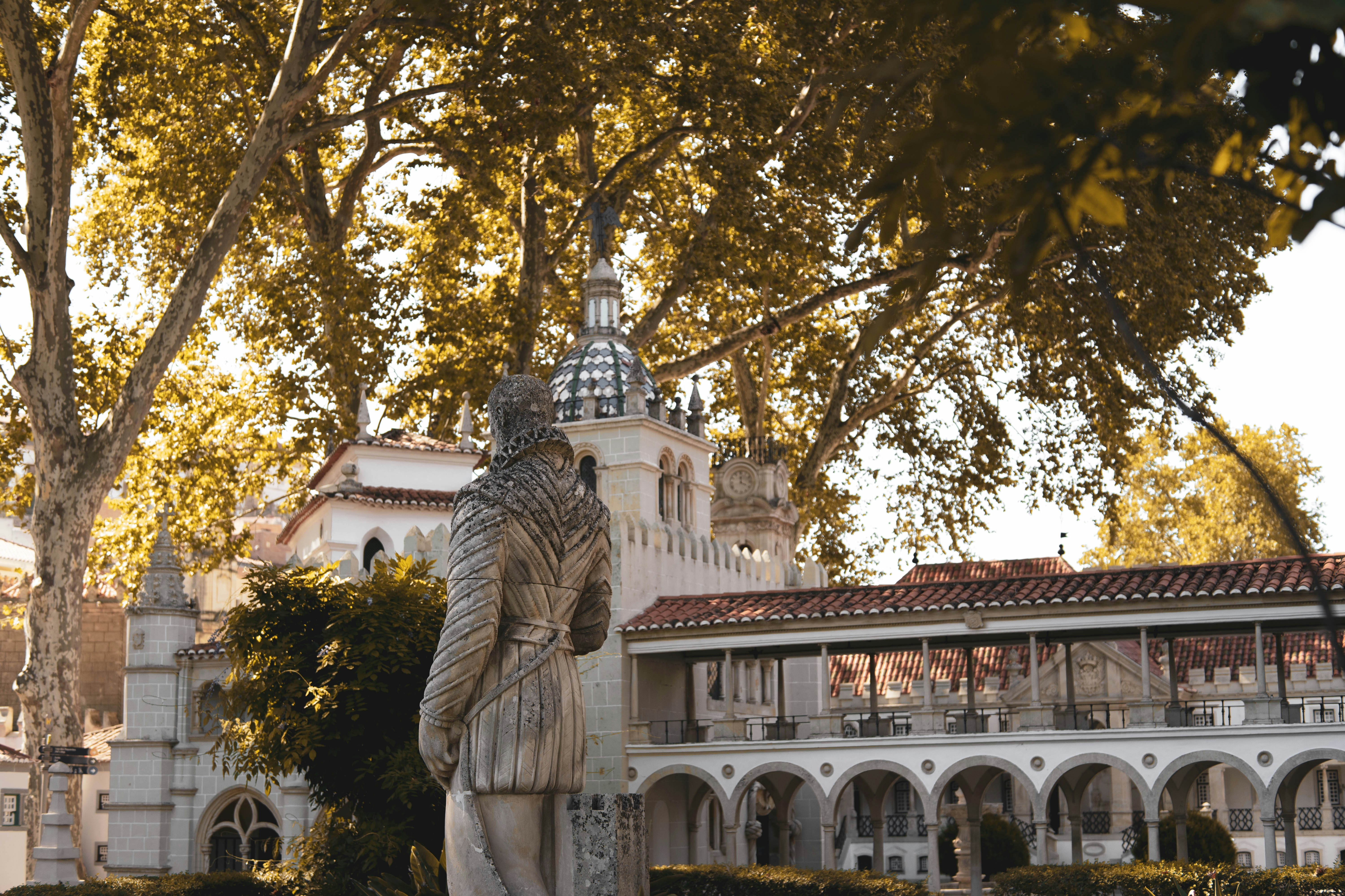 Stone statue beneath tall trees in front of a historic building with arched windows and a tiled roof.