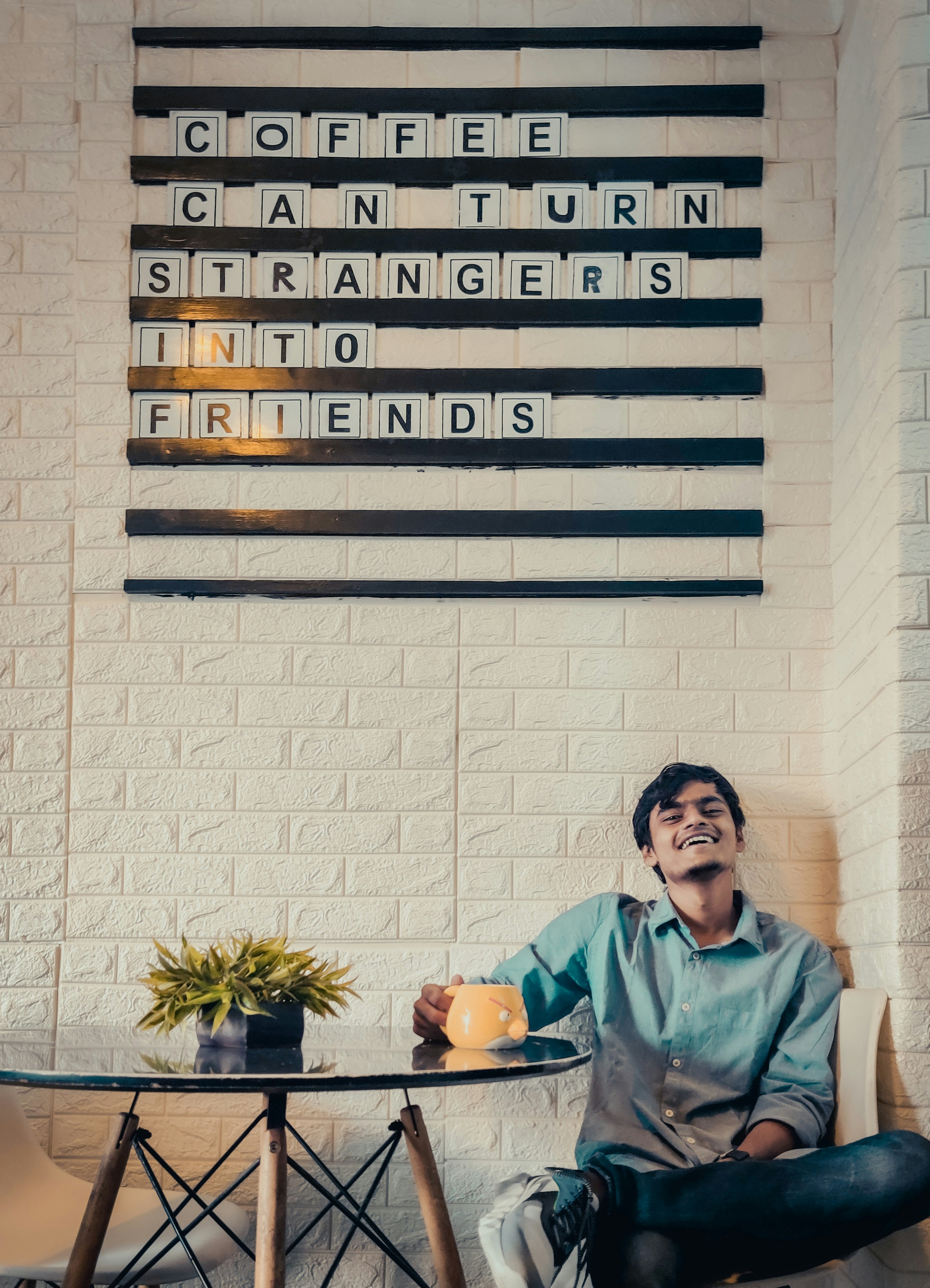 A young man enjoys a cup of coffee while seated at a table, with a playful wall display reading 'COFFEE CAN TURN STRANGERS INTO FRIENDS.'