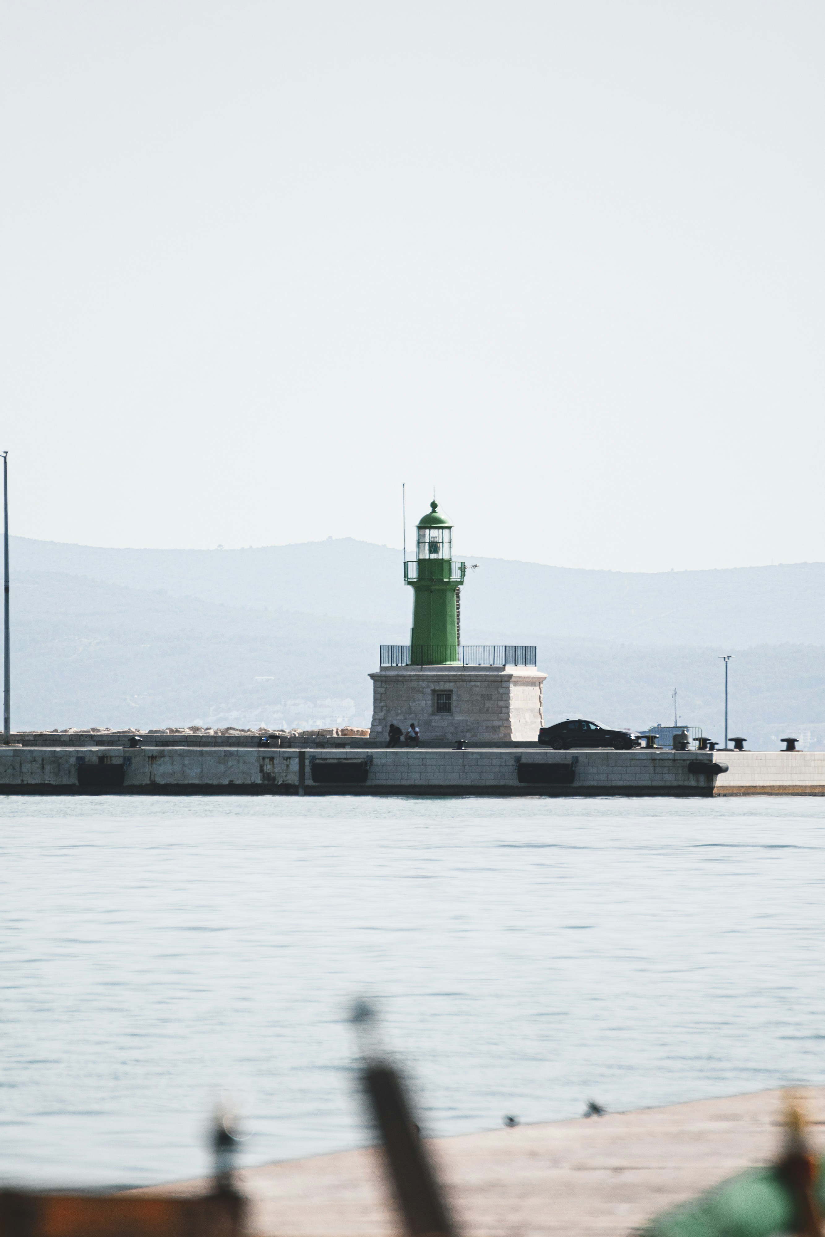 Green lighthouse standing on a pier with gentle waves reflecting its silhouette against a hazy backdrop of distant mountains.