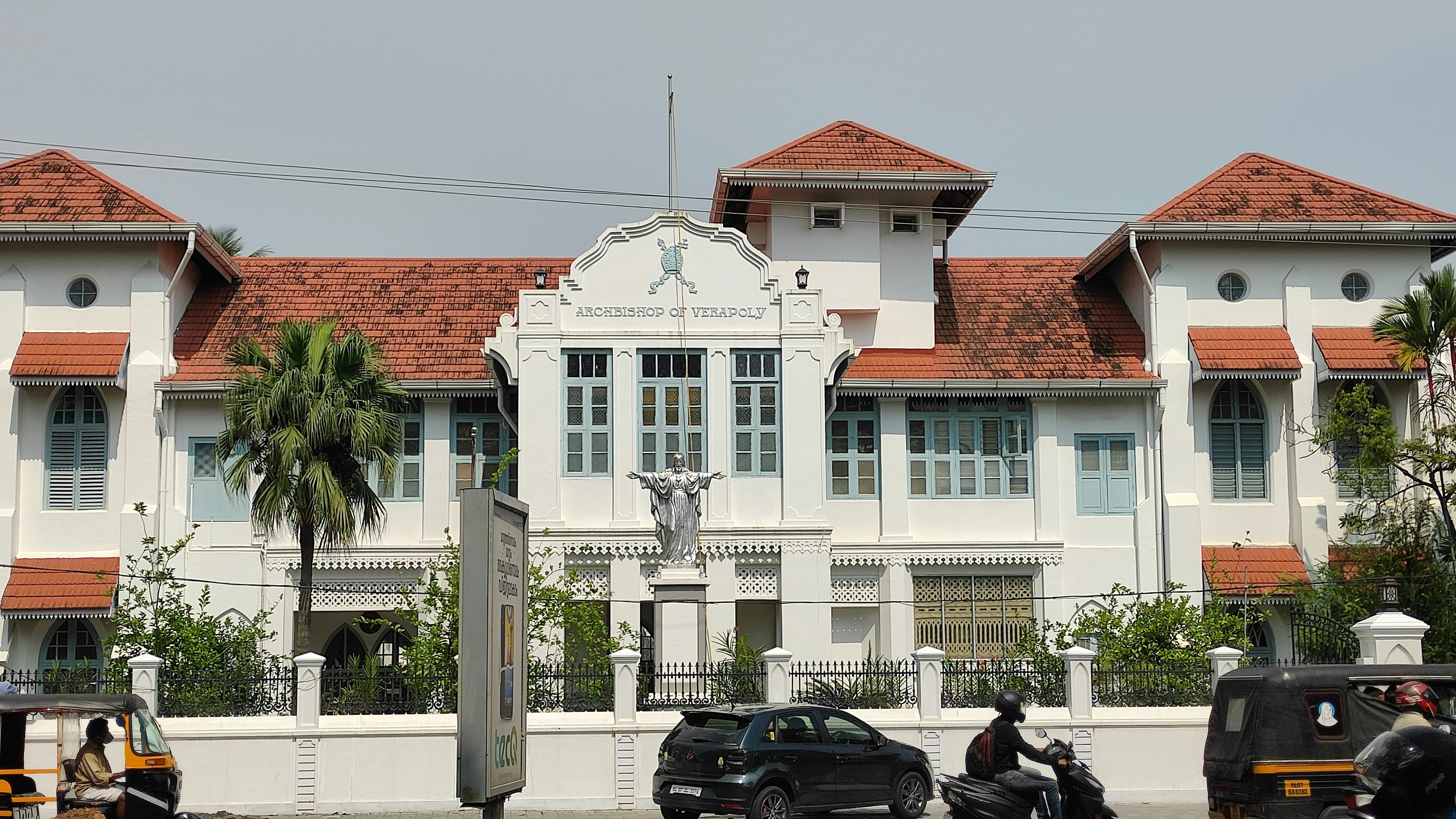 Elegant colonial-style building featuring intricate architectural details and a prominent statue, surrounded by lush greenery and urban life.