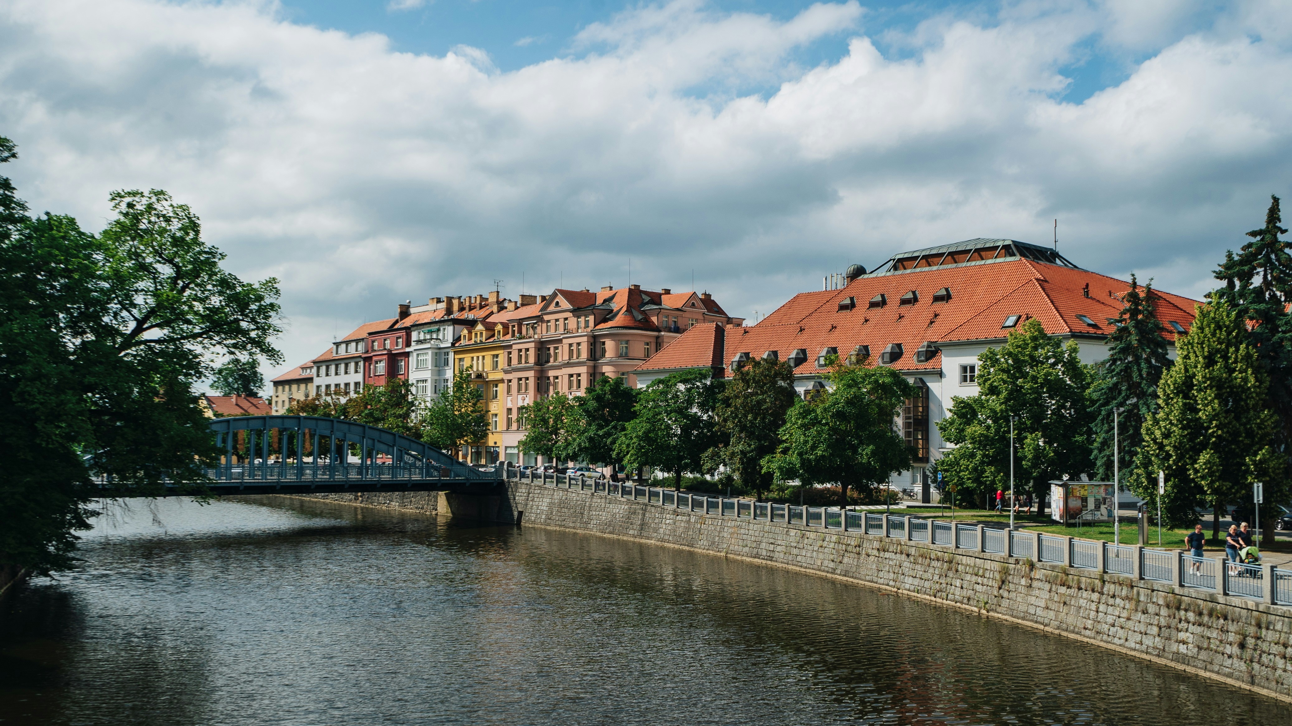 Historic buildings with red roofs line a tranquil river under a partly cloudy sky.