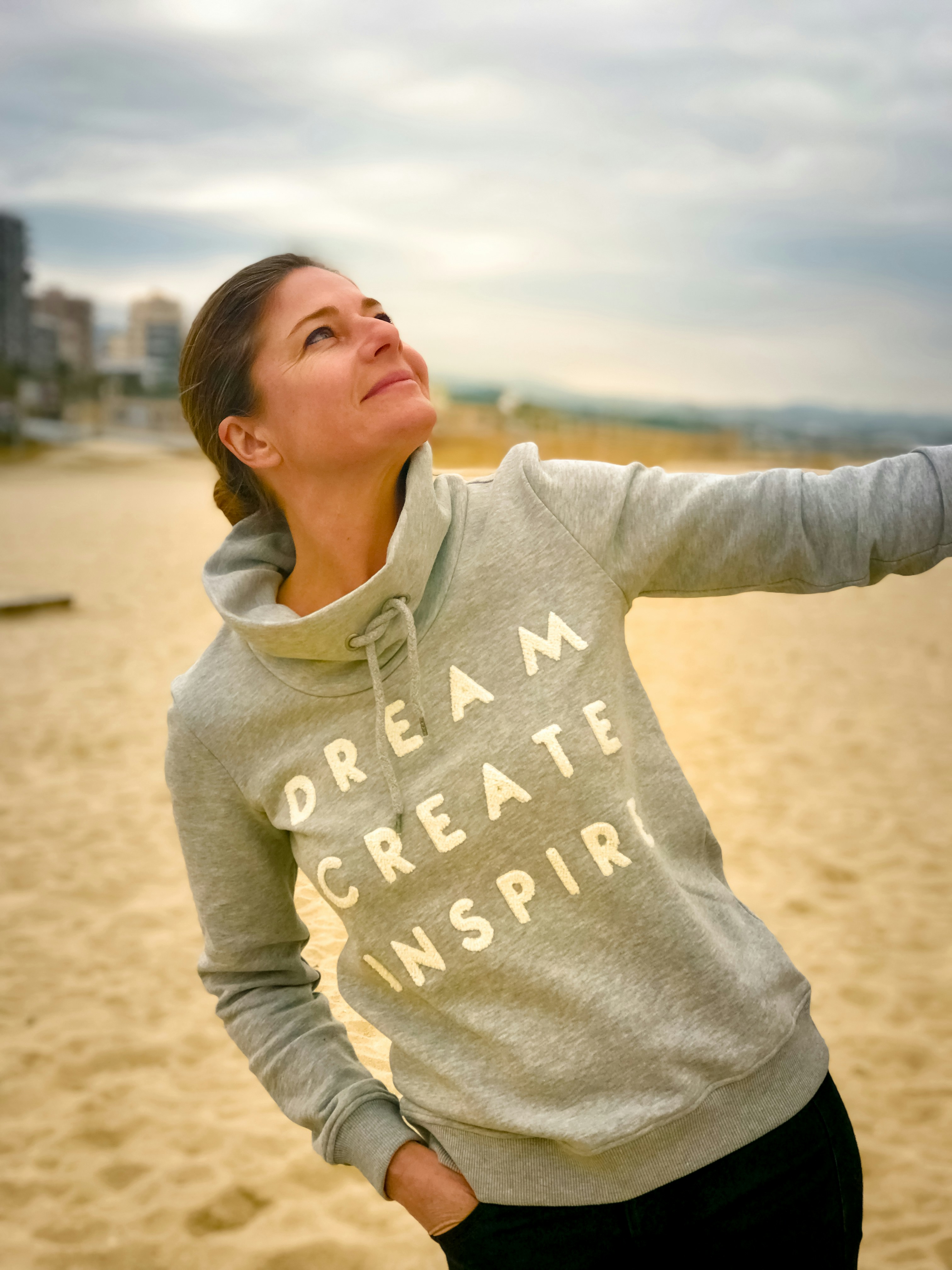 Woman in a gray hoodie with motivational text gazes thoughtfully into the distance on a sandy beach. The urban backdrop adds contrast to the serene setting.