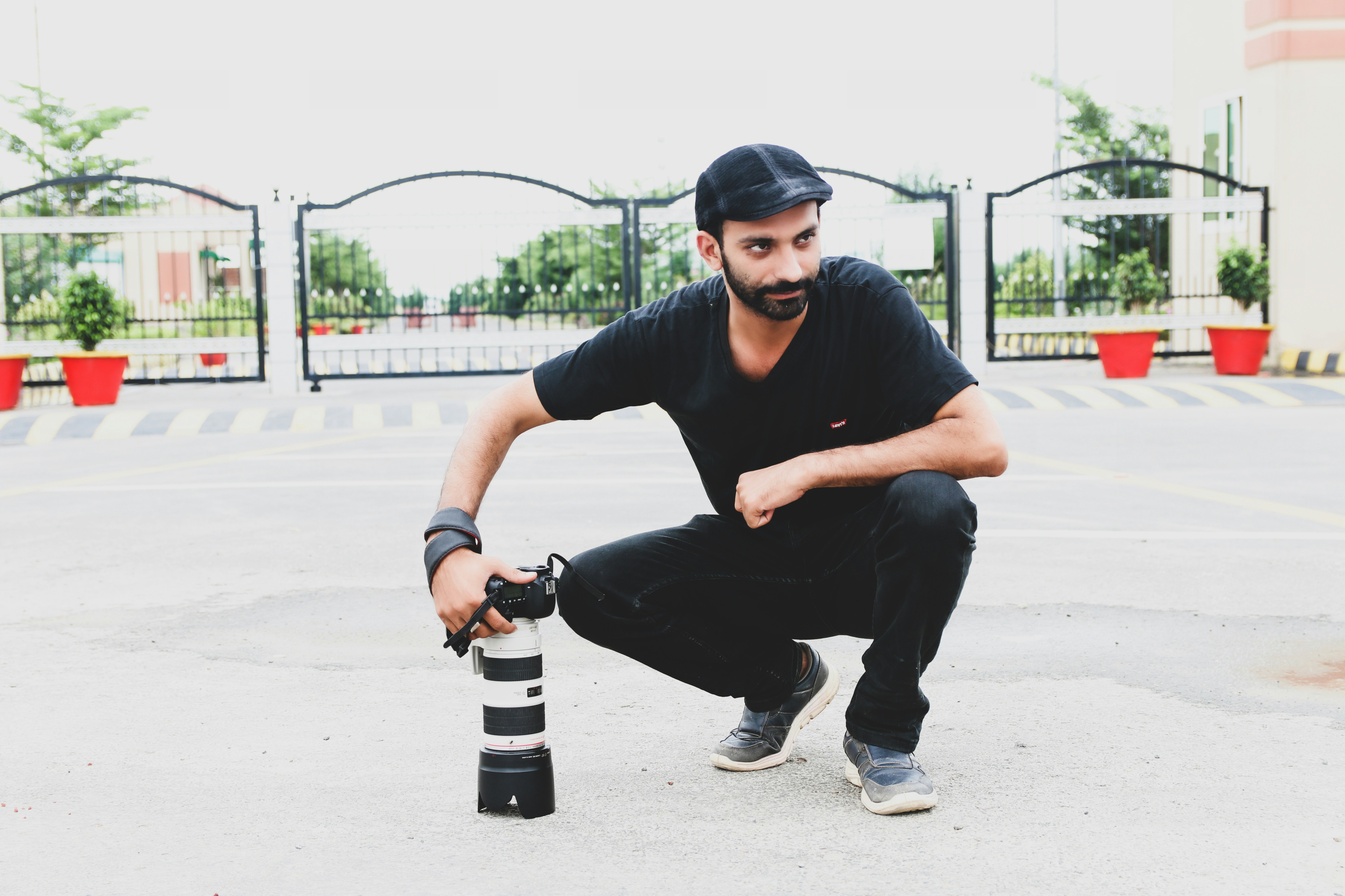 Man crouching on pavement holding a camera with a large lens, framed by a gated background and red potted plants.