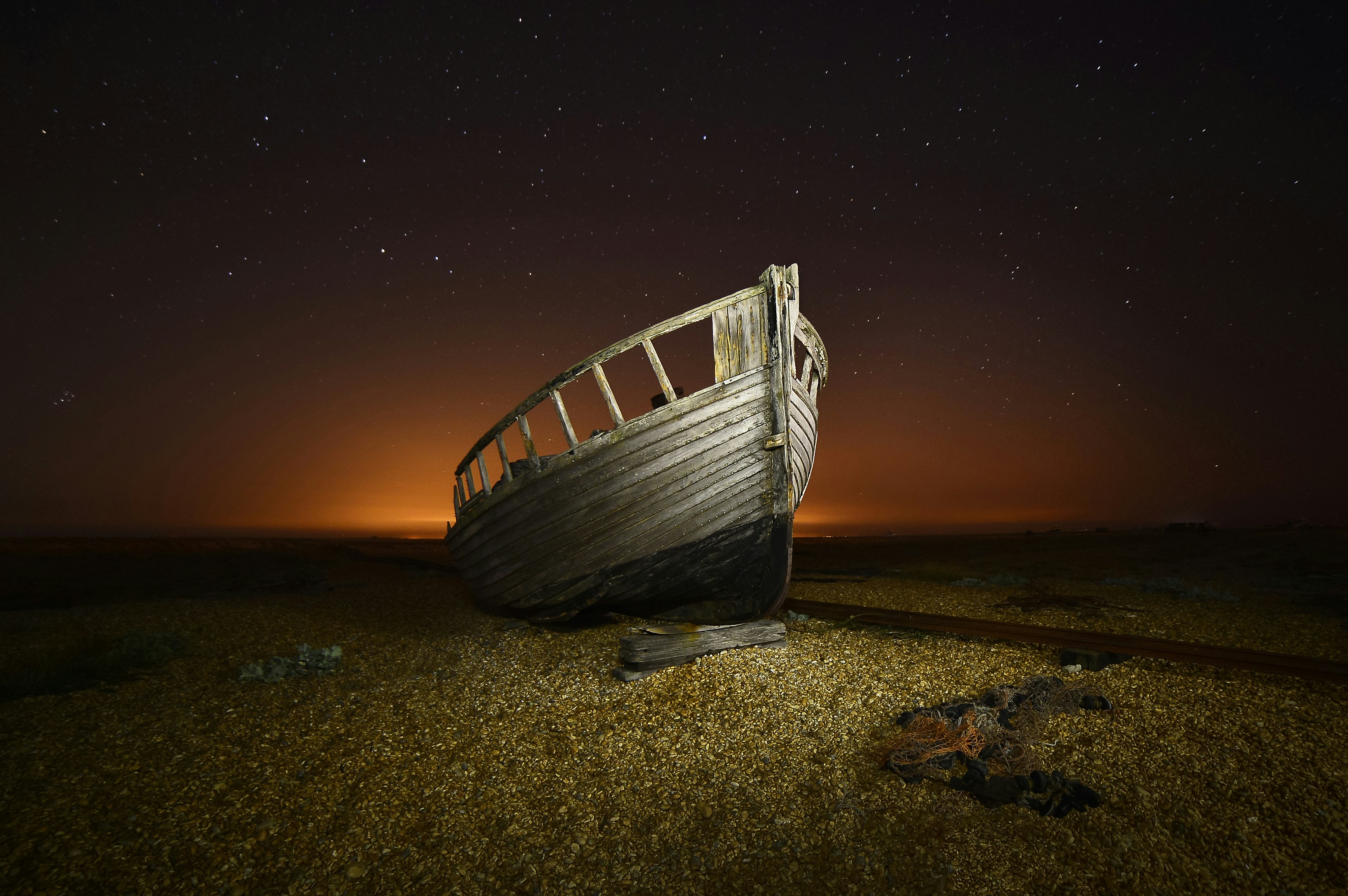 Old wooden boat illuminated under a starry night sky with a warm glow on the horizon.