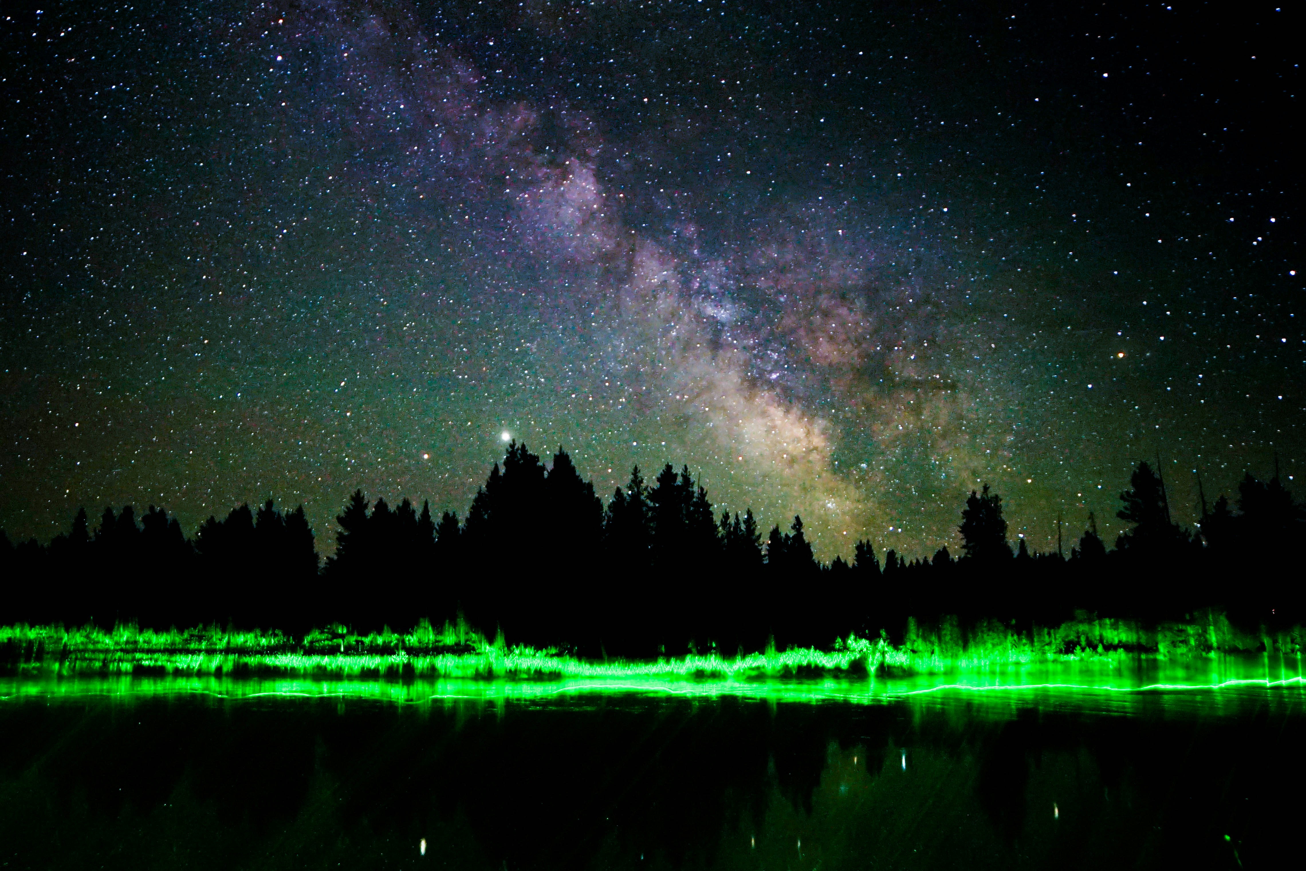 Milky Way galaxy arching over a serene lake, illuminated by vibrant green auroras reflecting on the water's surface.