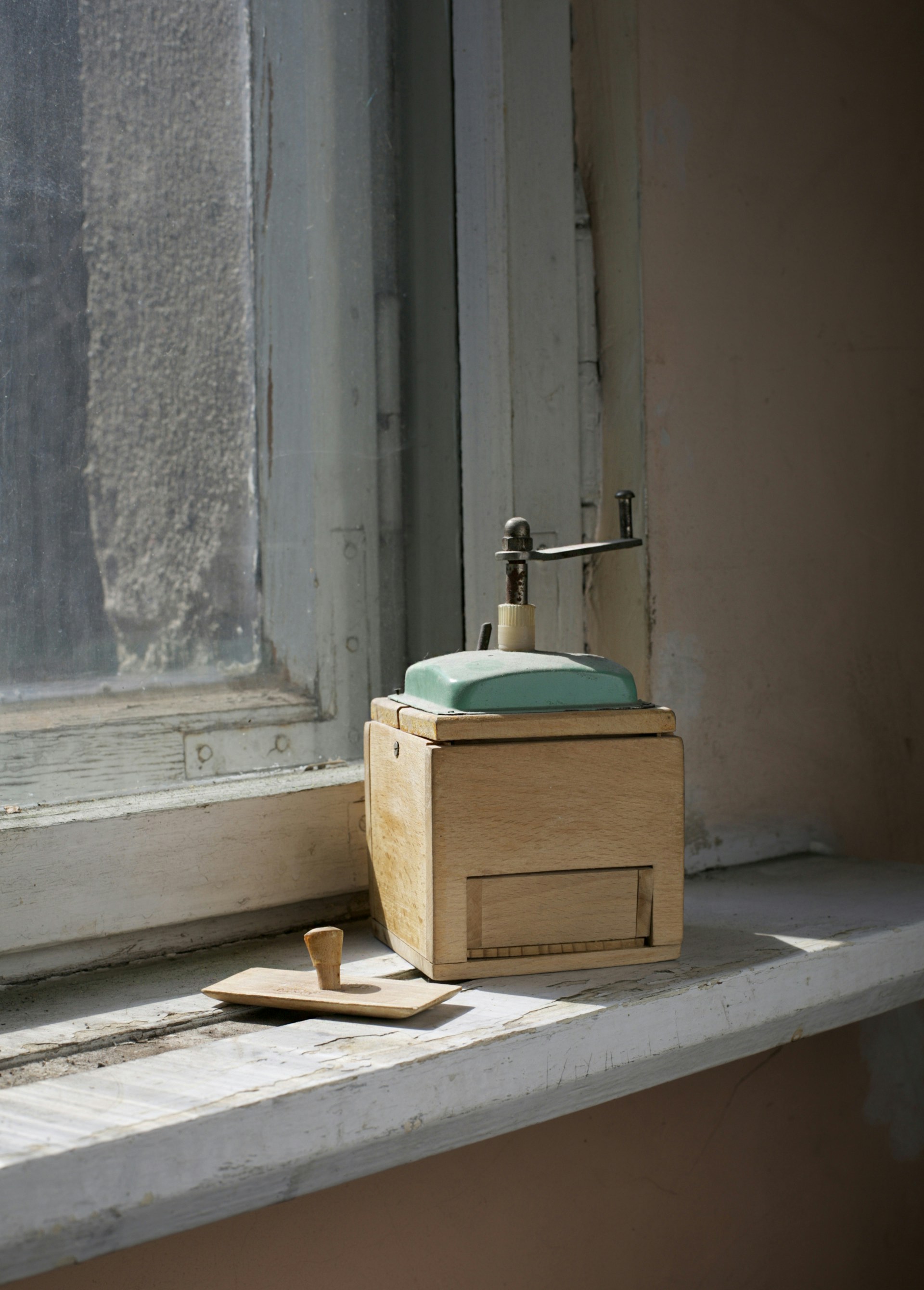 A vintage wooden coffee grinder sits on a sunlit windowsill, accompanied by a small wooden scoop. The soft light enhances the textures of the wood and the pastel green lid.