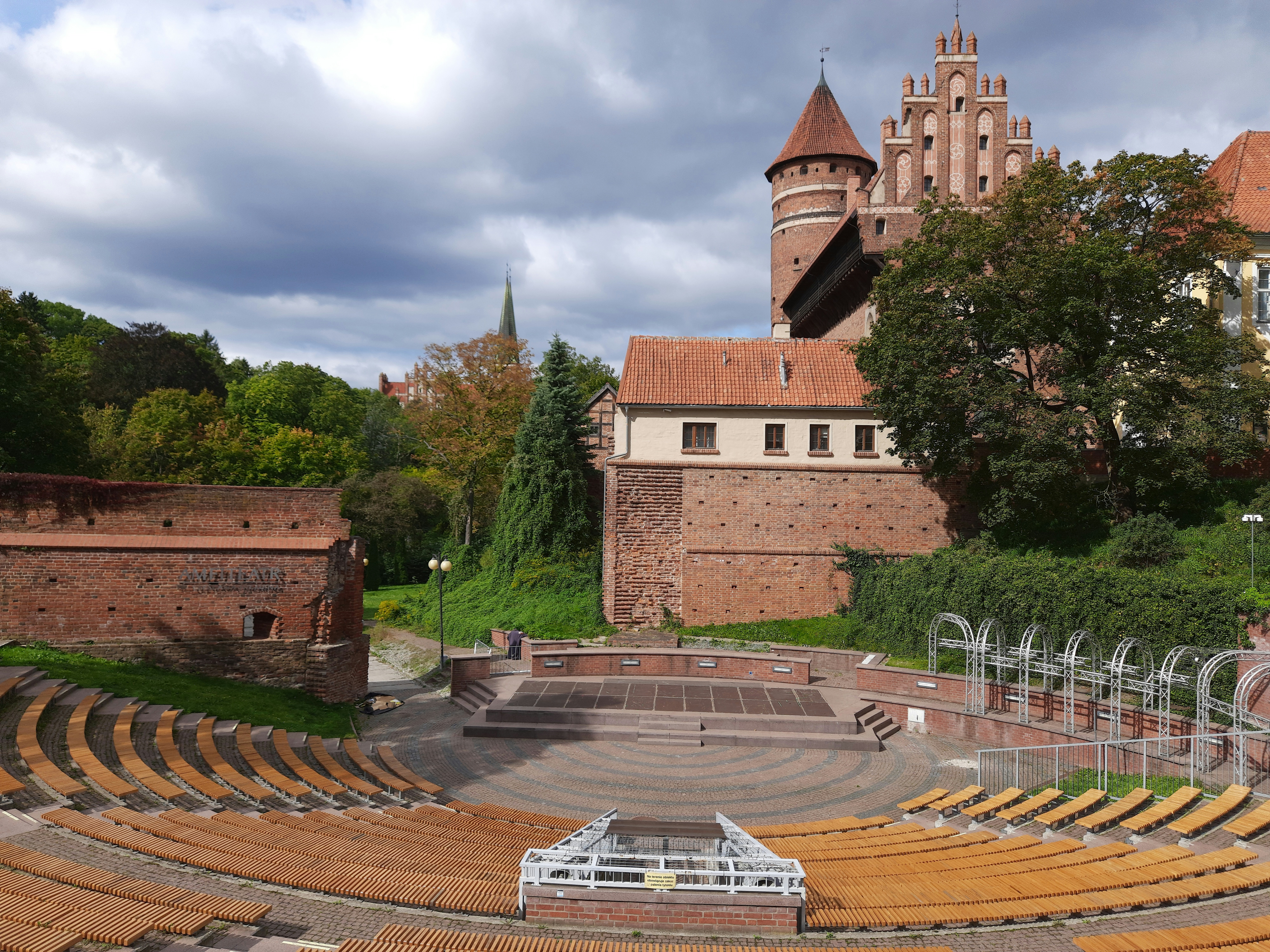 An open-air amphitheater nestled against a historic backdrop, featuring wooden seating and surrounded by lush greenery and ancient architecture.