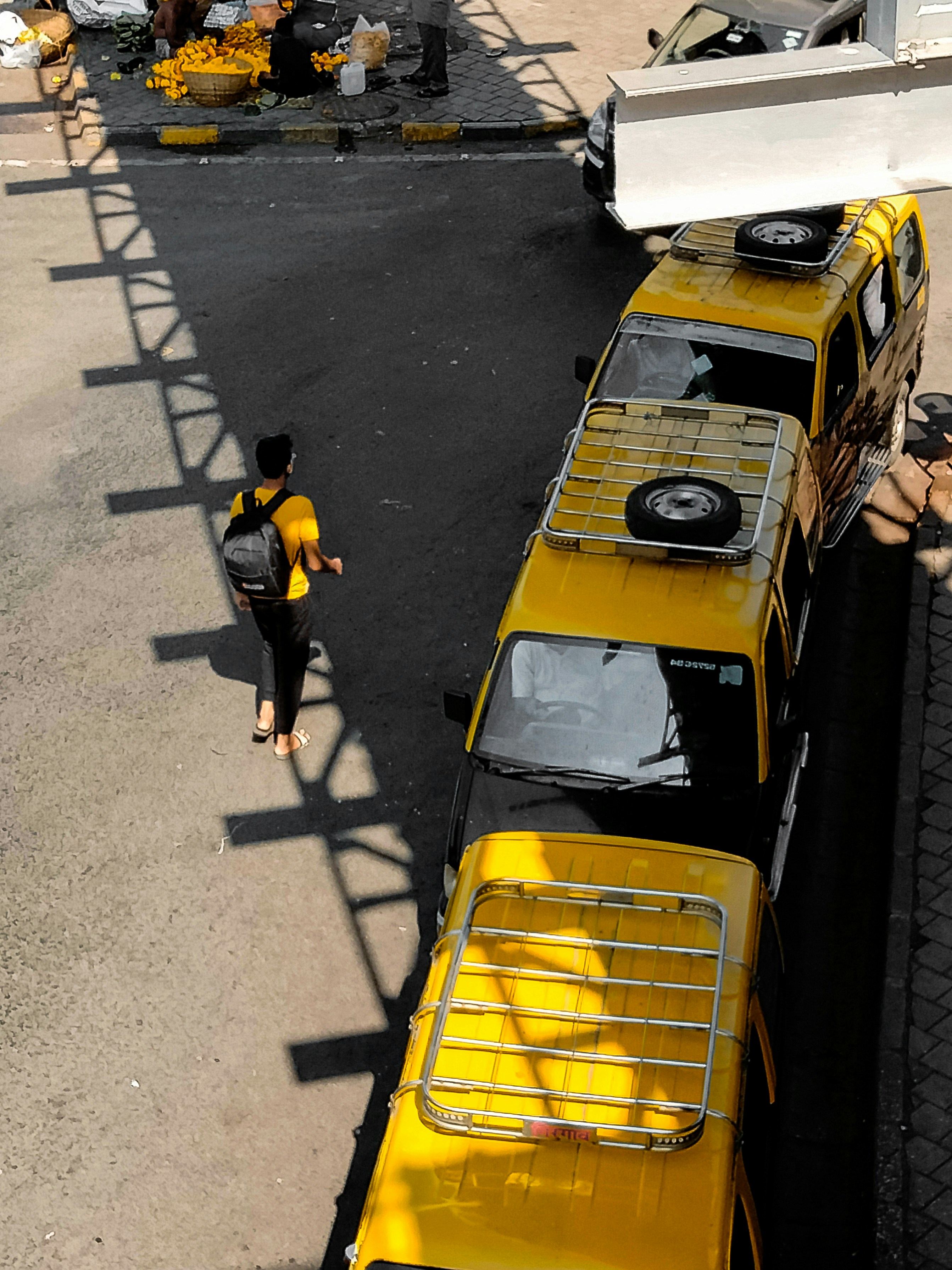 Top-down street view showing a row of yellow taxis along the curb and a pedestrian in a yellow shirt crossing the frame. Strong shadows from a scaffold create bold geometric patterns.
