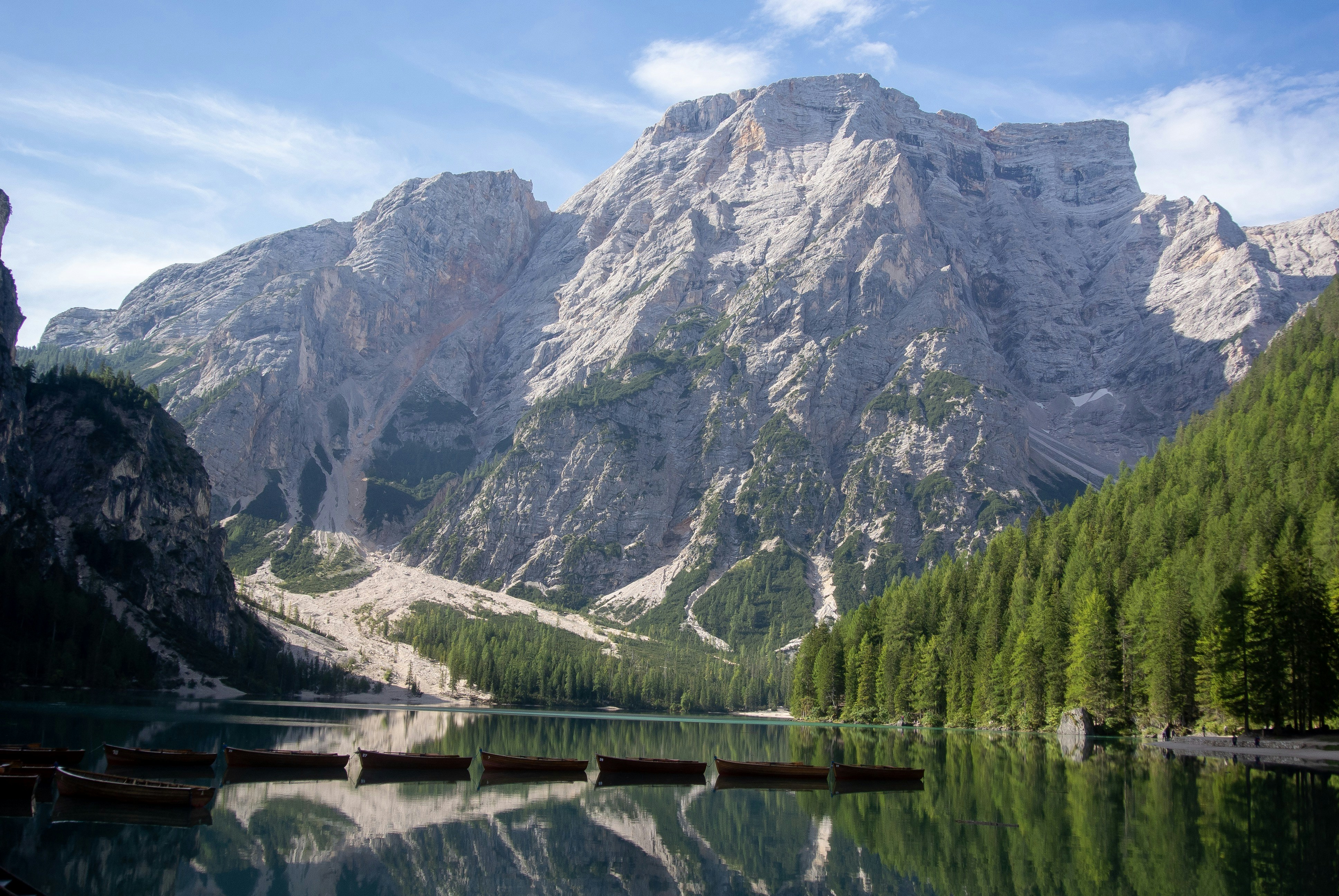 Serene lake reflecting towering Dolomite mountains under a clear sky, framed by lush green forests and wooden boats at rest.