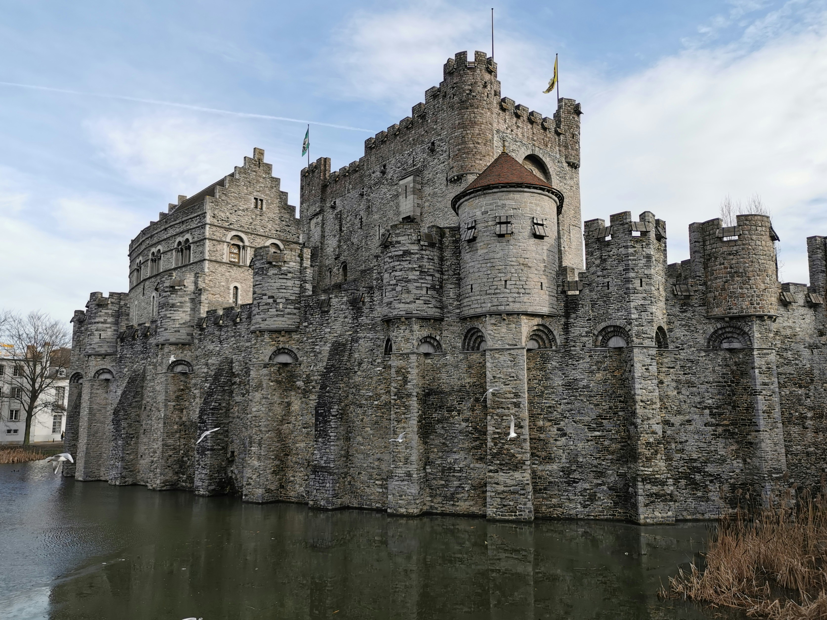 Stone castle with turrets and battlements beside a calm moat under a partly cloudy sky.