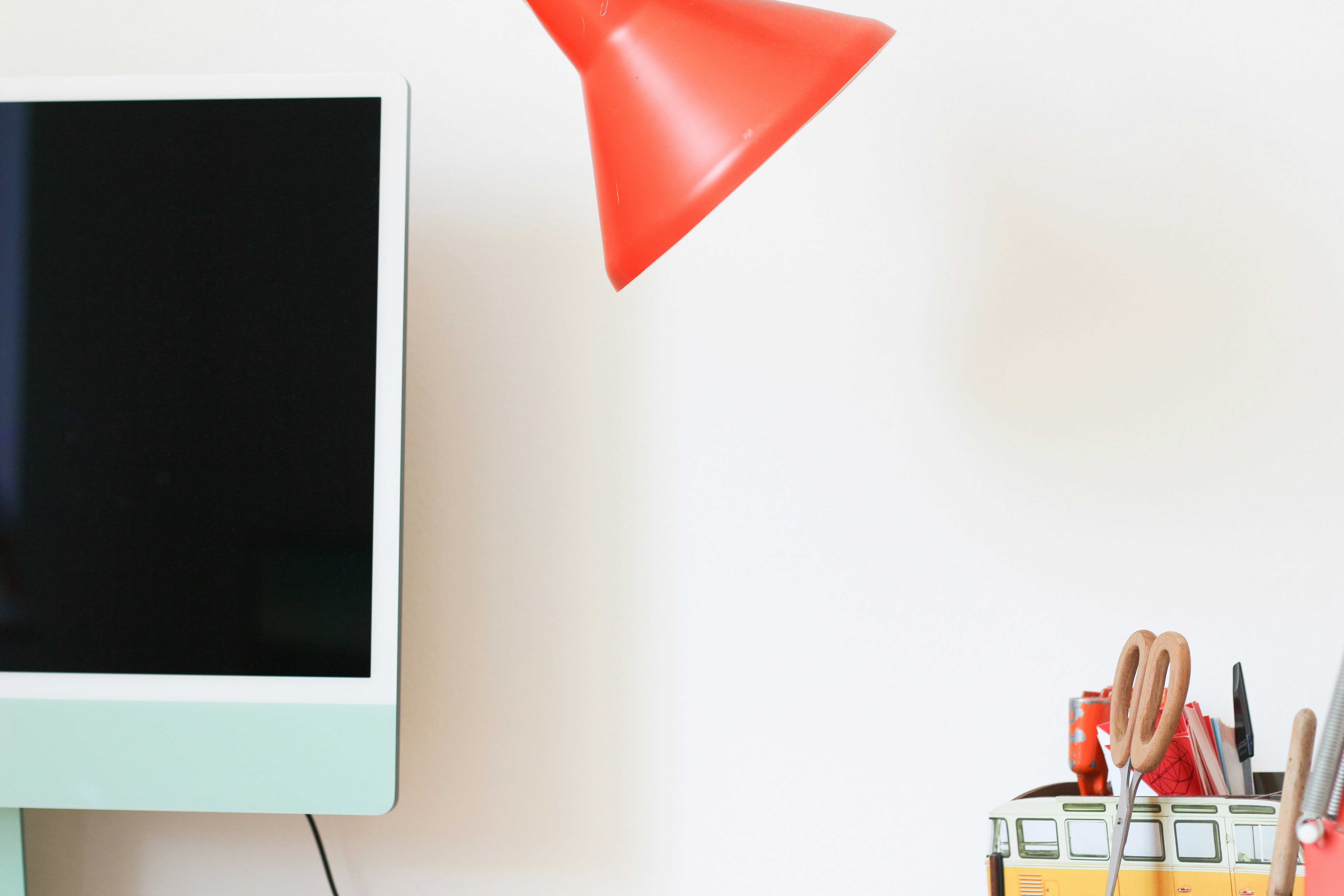Bright red lamp casts a warm glow over a stylish workspace featuring a vintage bus-shaped pencil holder and an oversized monitor.