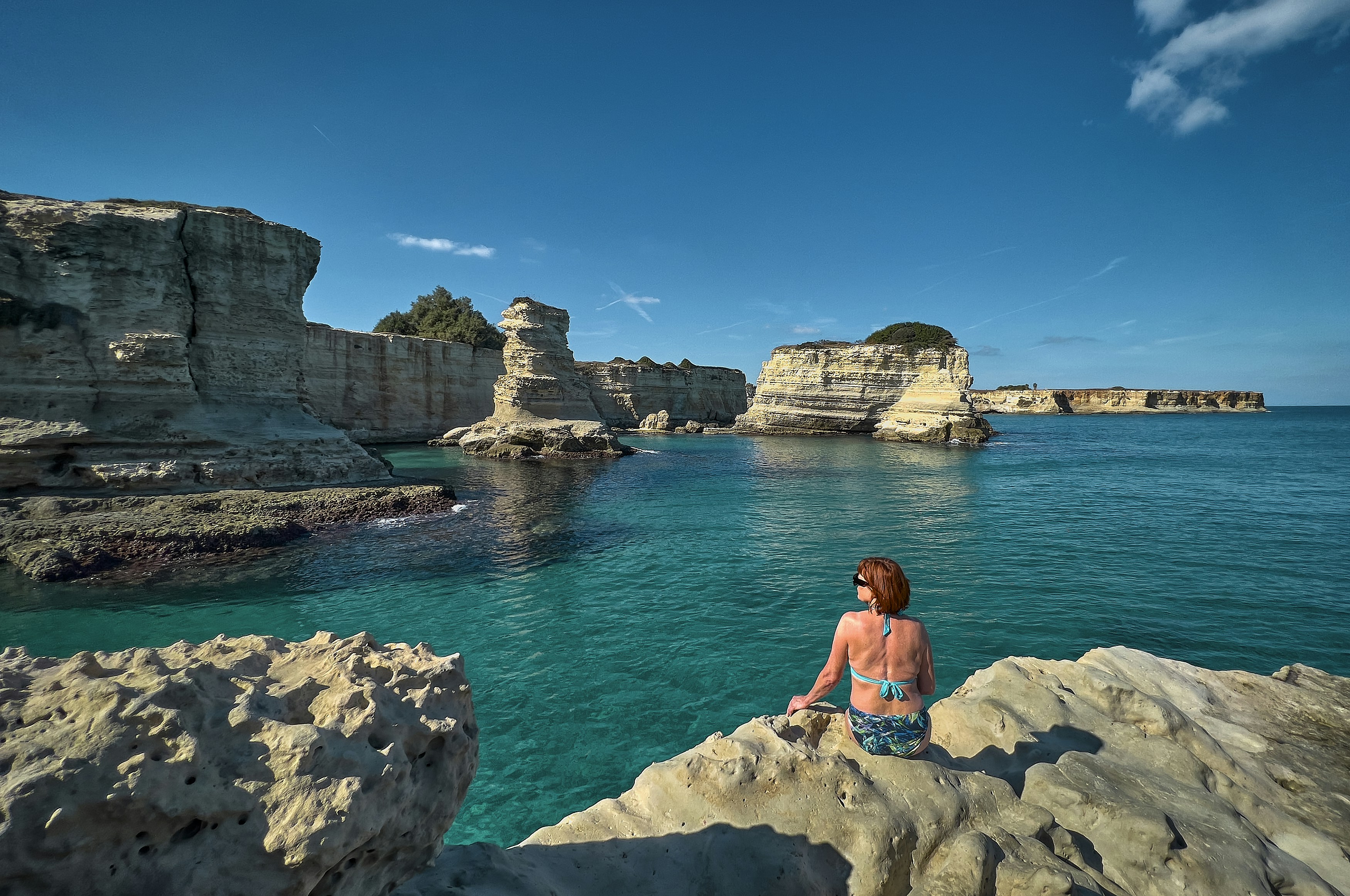 Person seated on a rocky outcrop overlooking turquoise ocean waters under a clear blue sky.
