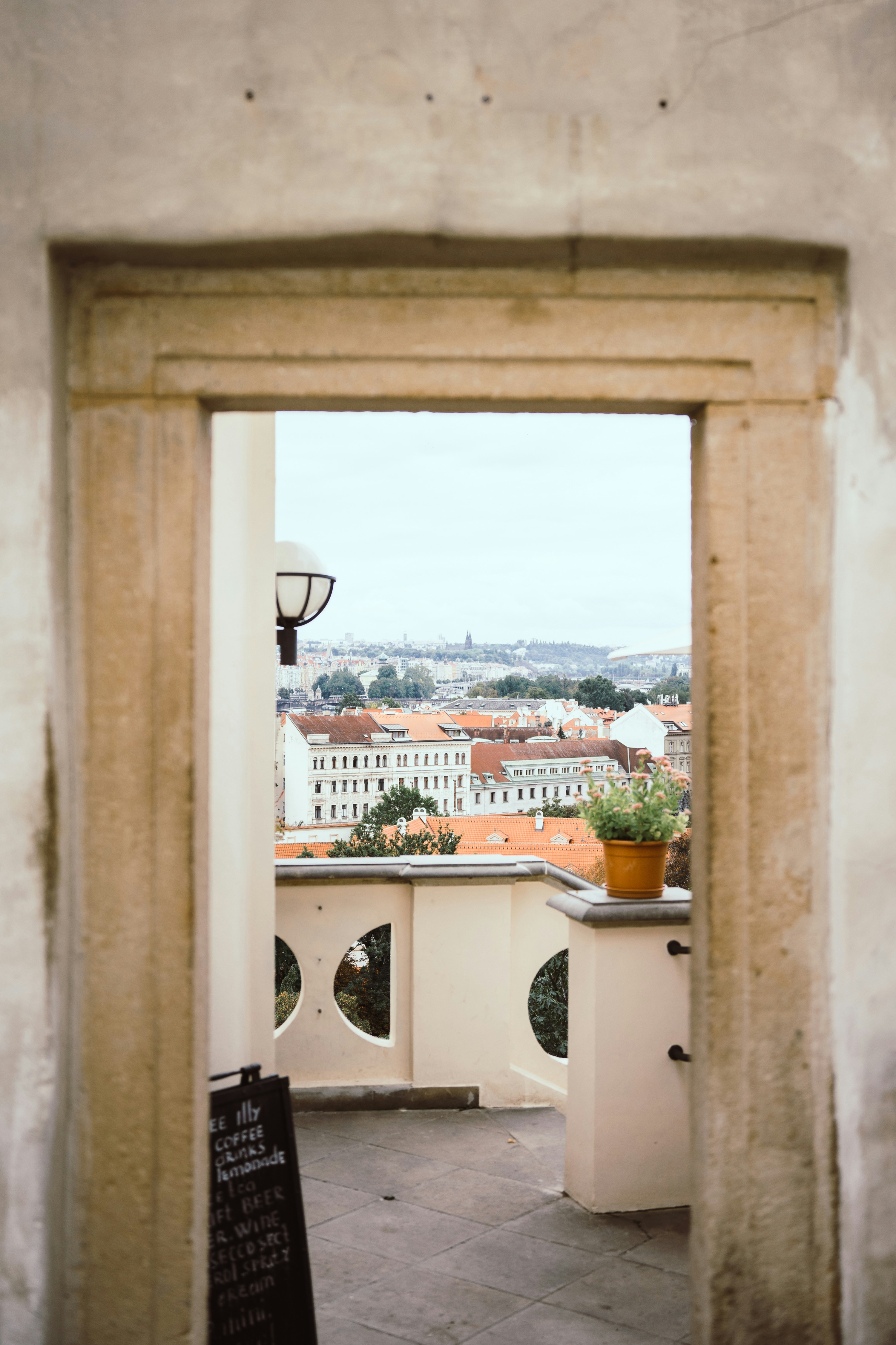 Open doorway revealing a picturesque view of a cityscape with terracotta rooftops and distant hills. A potted plant adds a touch of greenery to the scene.