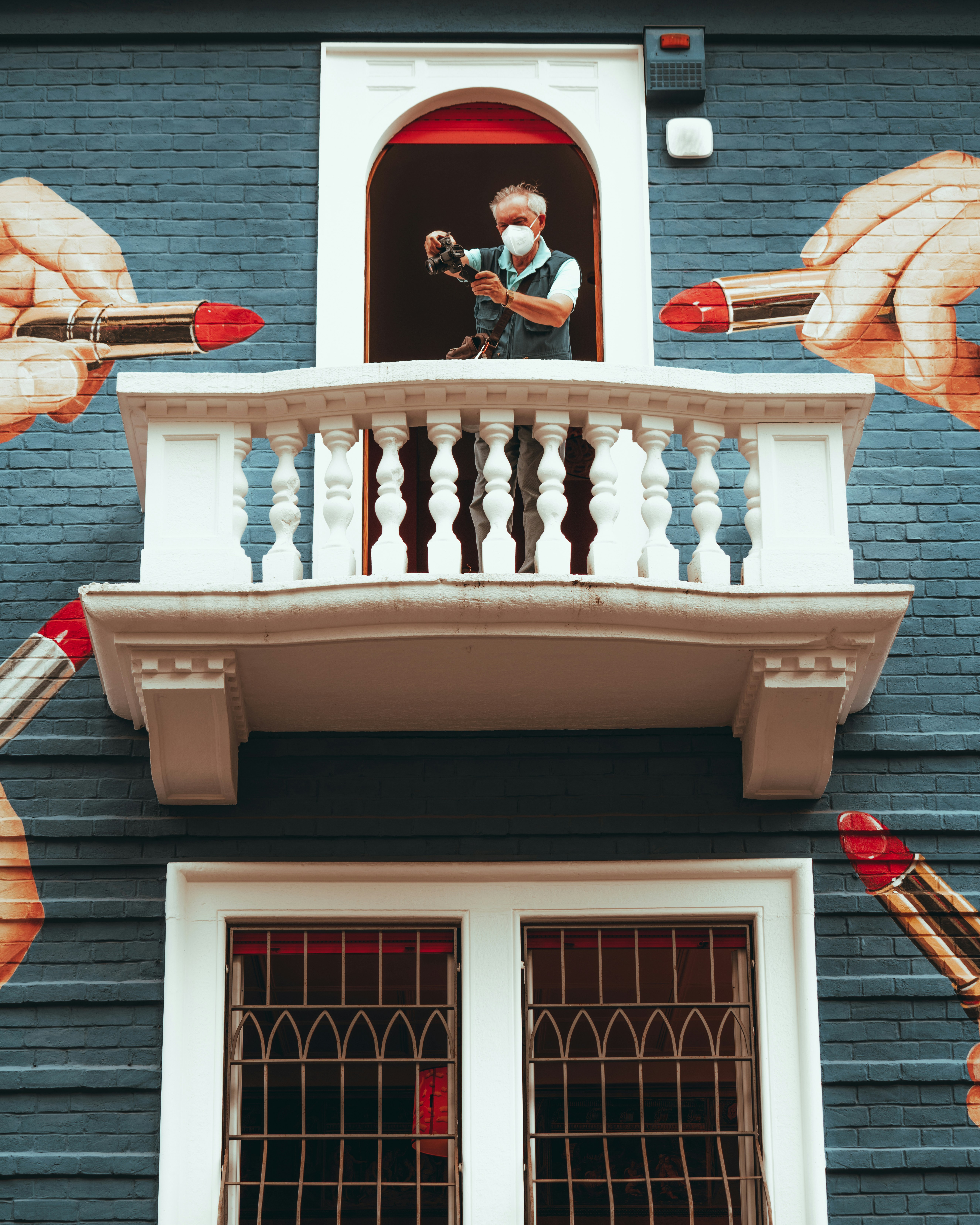 A person stands on a balcony, holding a camera, with vibrant lipstick murals framing the scene. The artwork celebrates creativity and self-expression.