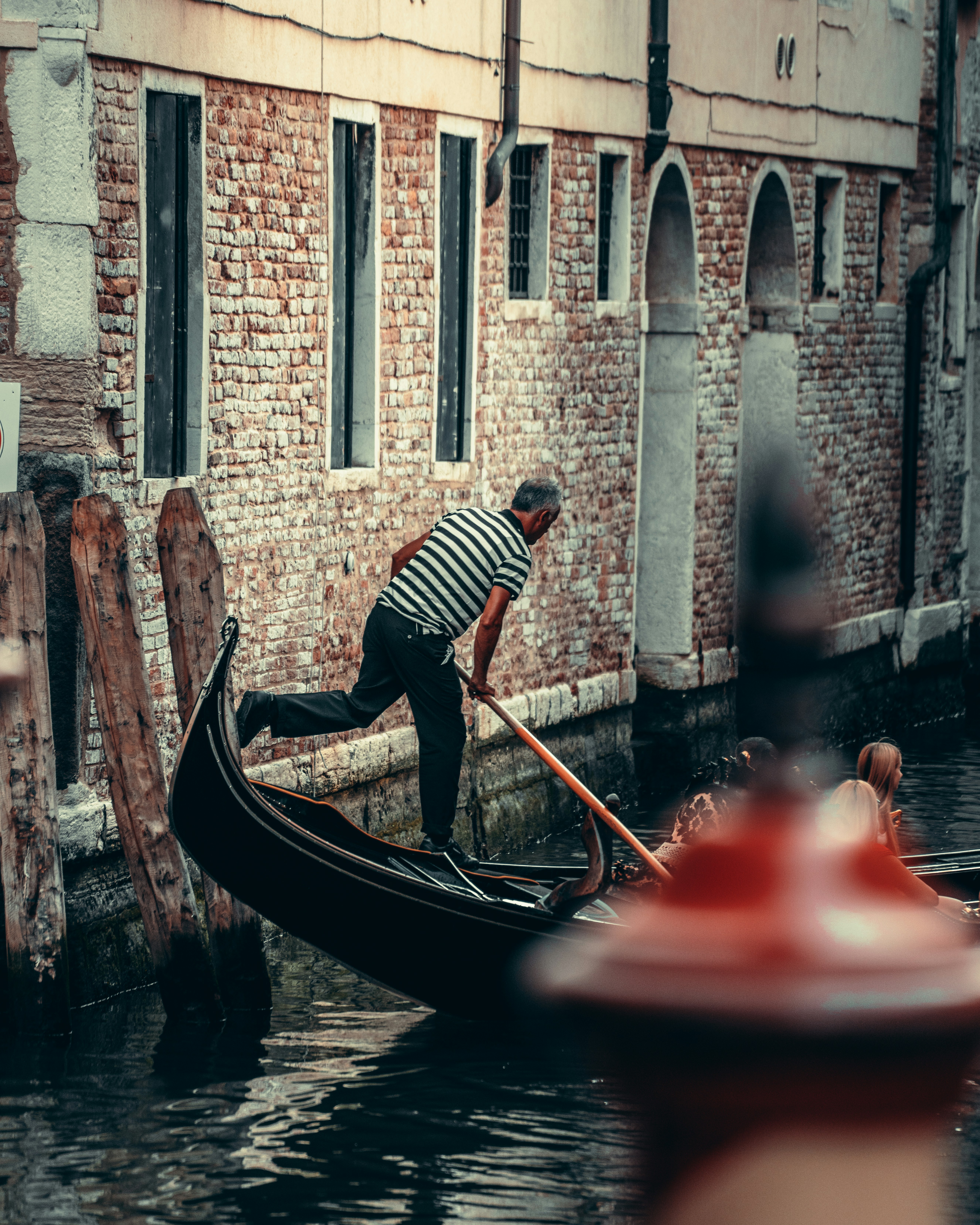 Gondolier in striped shirt maneuvering a gondola through a narrow Venetian canal, with historic brick buildings in the background.