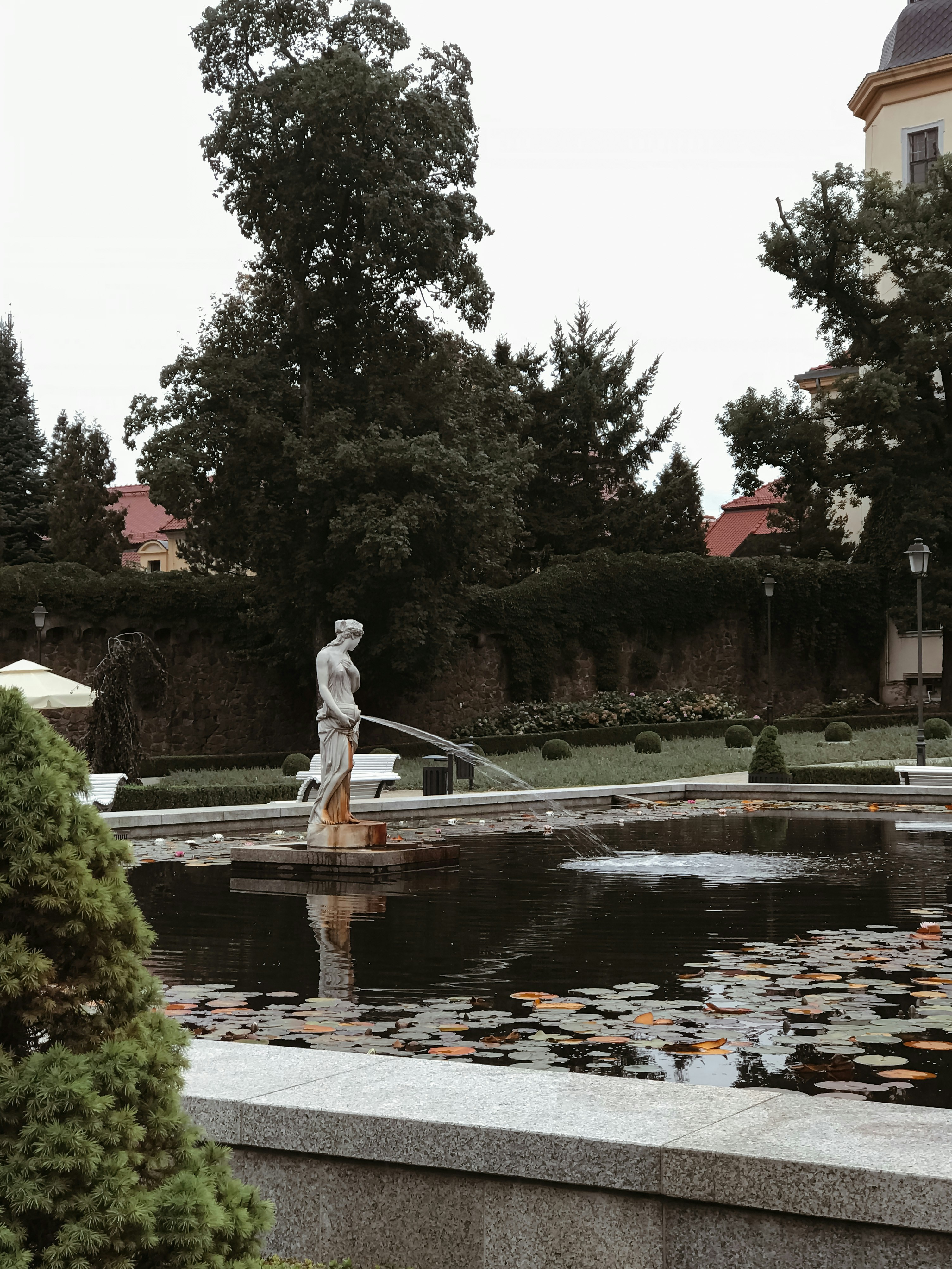 A classic statue of a figure pouring water stands at the edge of a tranquil pond surrounded by manicured greenery and lily pads.