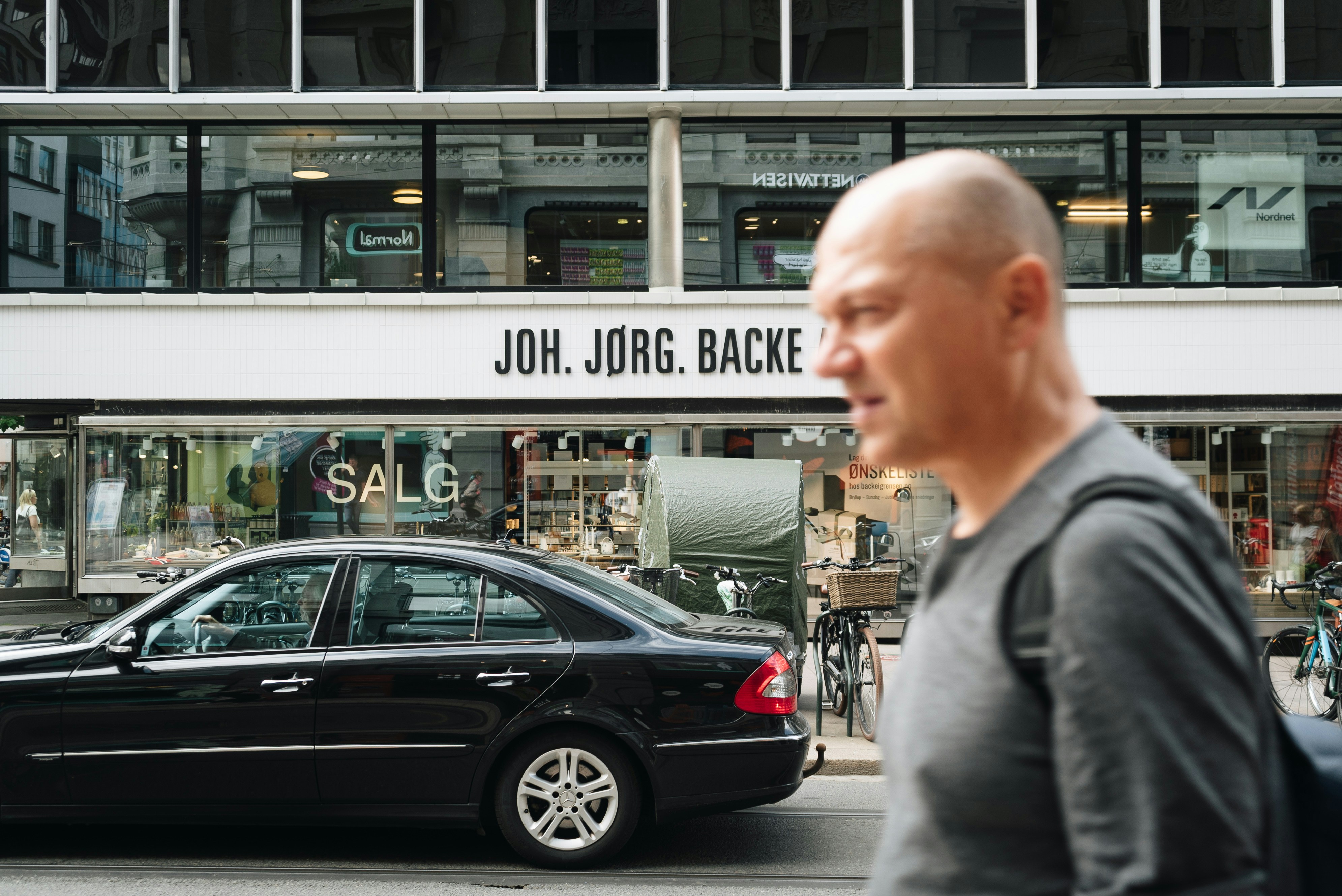 A blurred figure walks past a sleek black car and a storefront, capturing the essence of city life in motion.