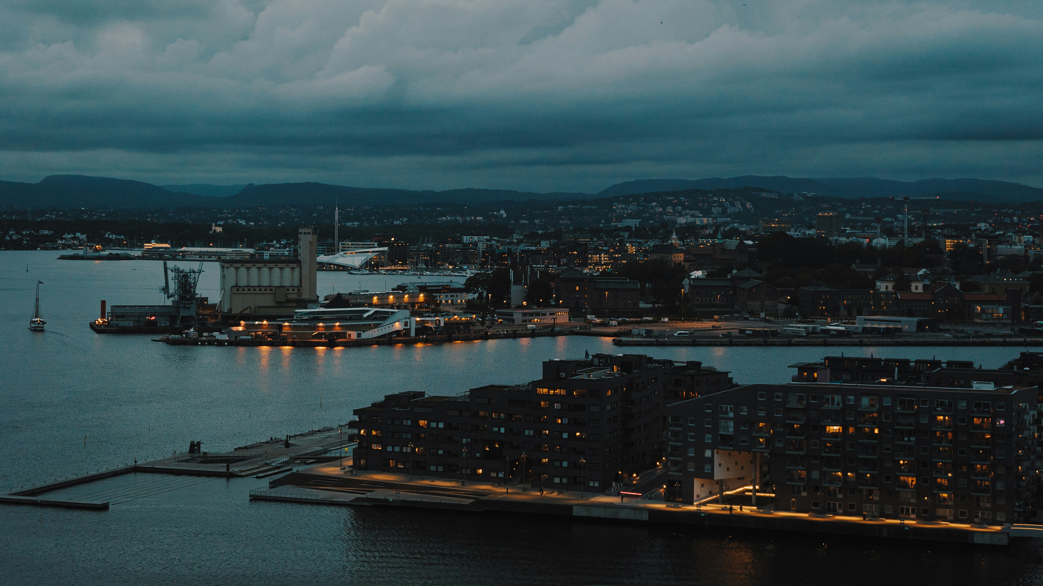 Cityscape at dusk featuring illuminated buildings along the waterfront and a harbor with boats. The scene captures the serene ambiance of an urban evening.