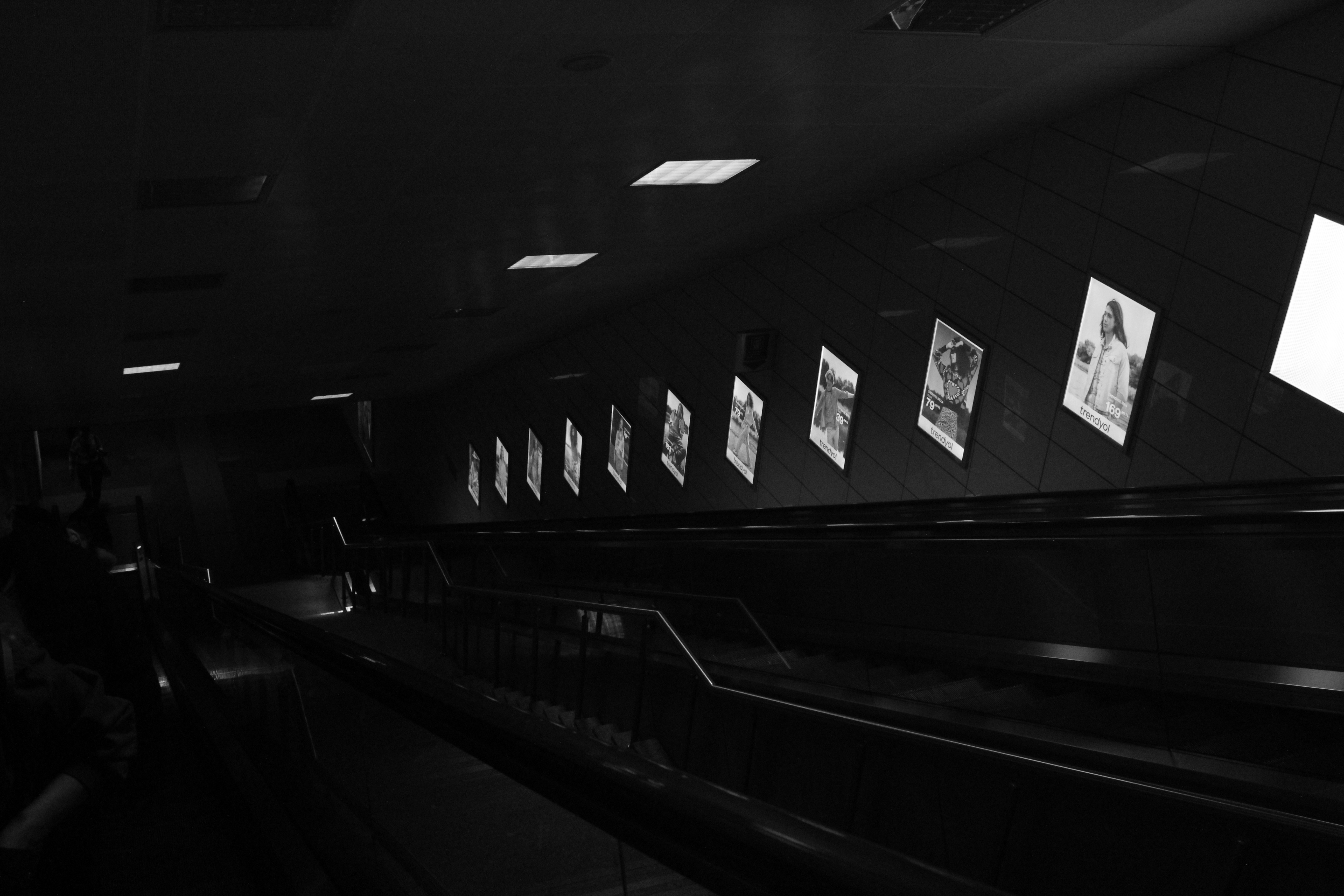 Black and white photograph showcasing an escalator lined with illuminated art displays in a subway station. The angle emphasizes the depth and perspective of the scene.