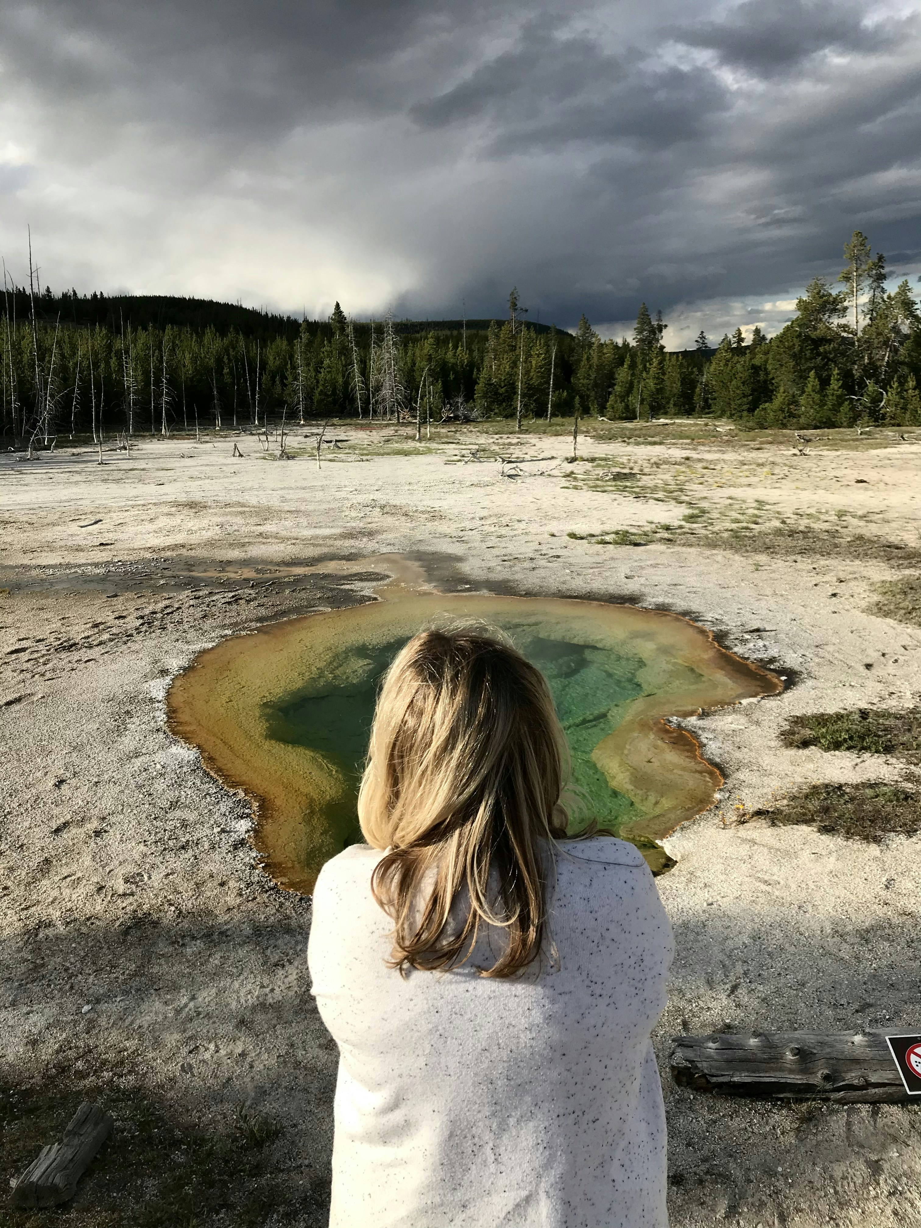 A woman gazes at a vibrant geothermal pool surrounded by a dry landscape and distant trees, capturing the contrast of colors and textures. The scene evokes a sense of tranquility amidst nature's wonders.
