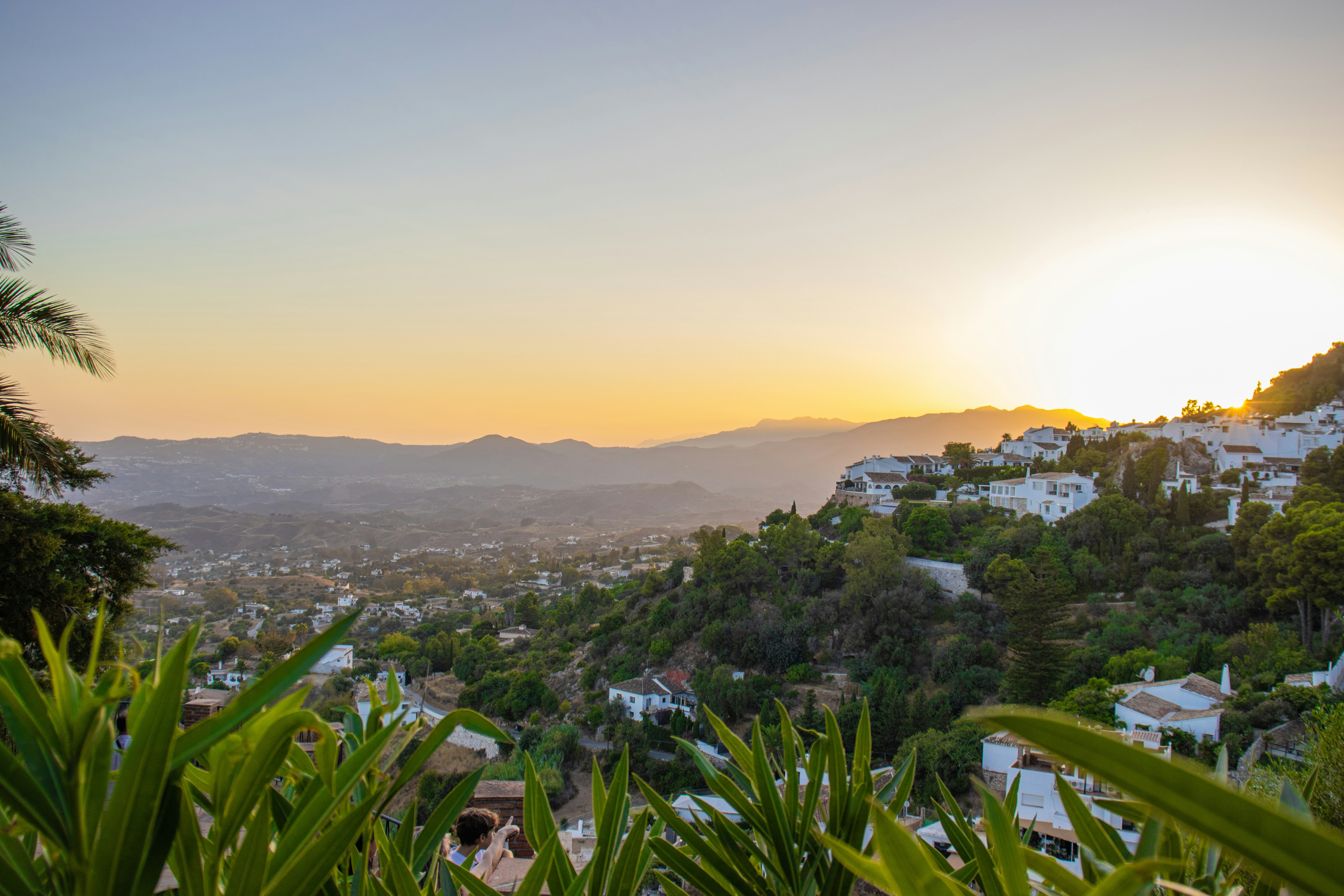 Sunset casting warm light over a hillside town with lush greenery and distant mountains.